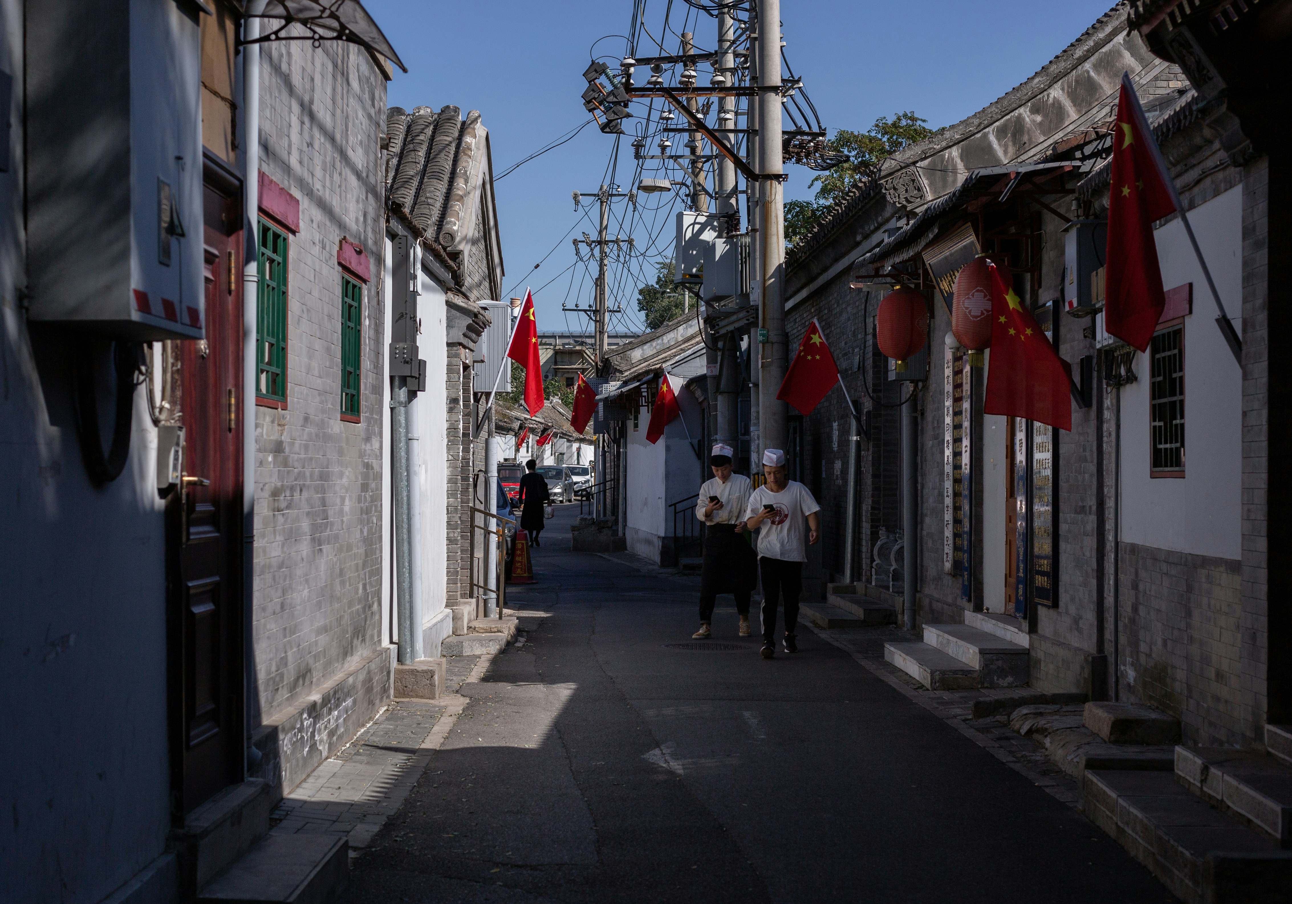 Narrow alleyway lined with traditional Chinese architecture, adorned with red flags, as two pedestrians stroll by under a clear blue sky.