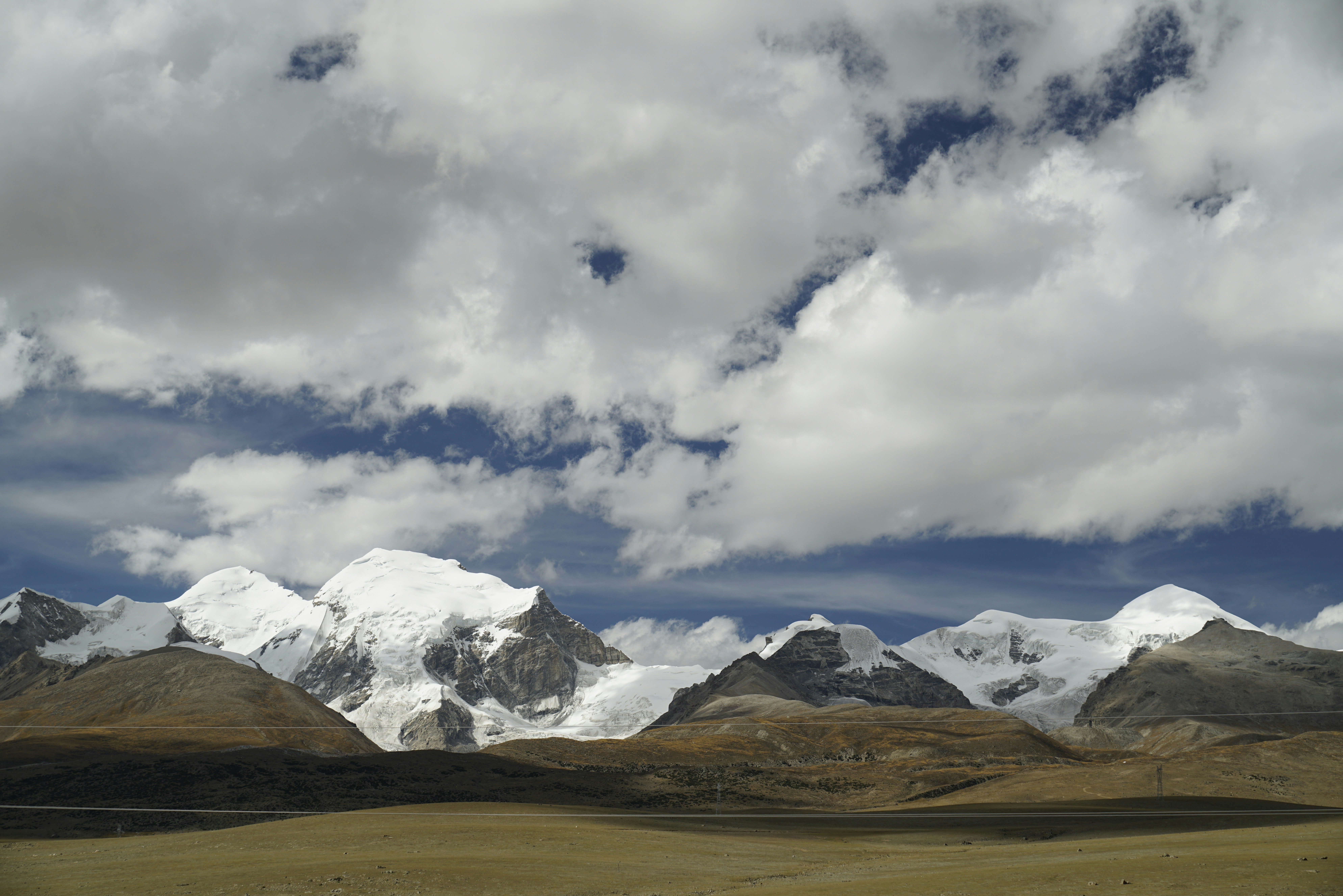 Snow-capped mountains rise dramatically against a backdrop of swirling clouds and blue sky. The vast landscape conveys a sense of tranquility and grandeur.