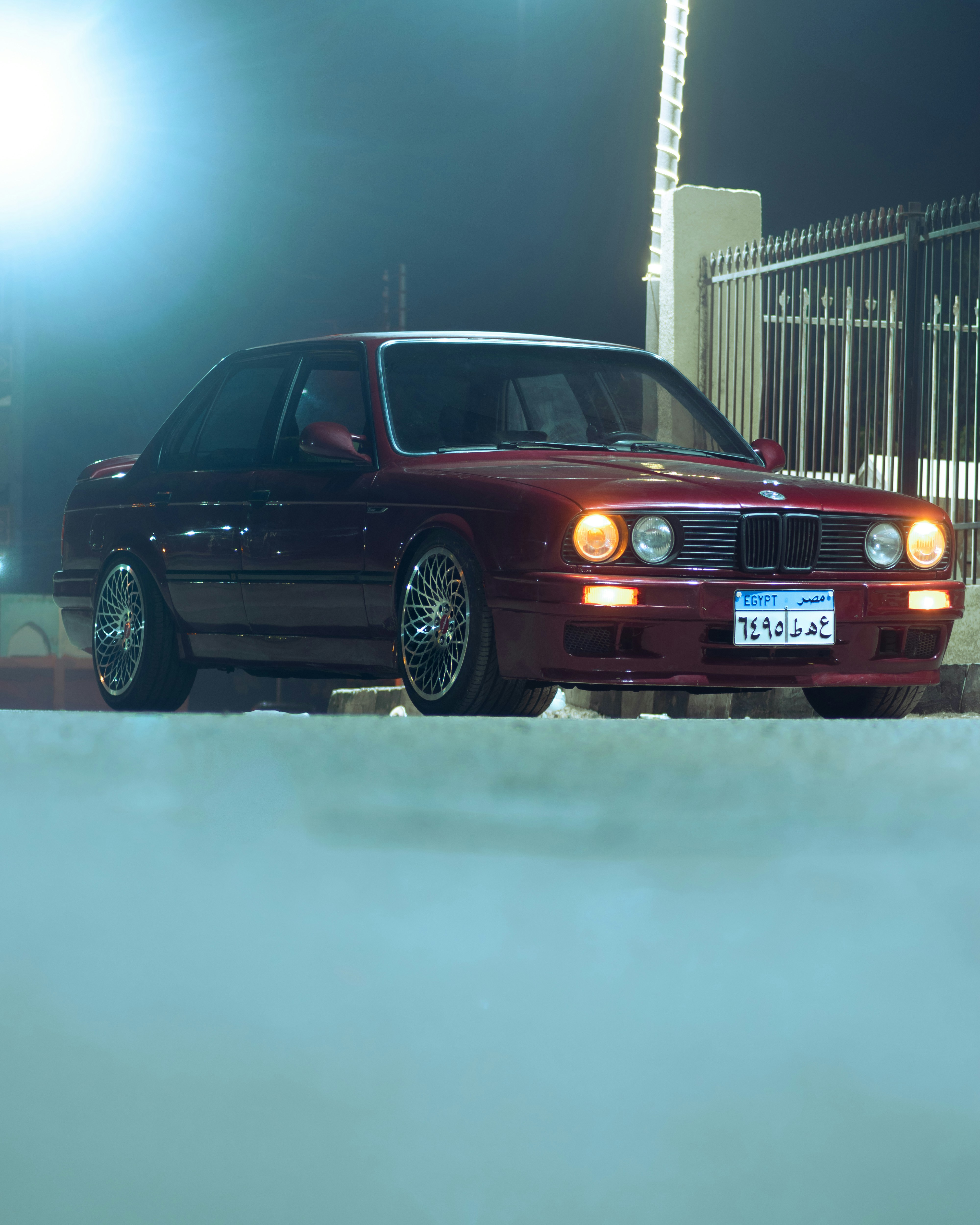 A classic red car parked under a night sky, illuminated by ambient lights. The vehicle showcases intricate wheel designs and a sleek profile.