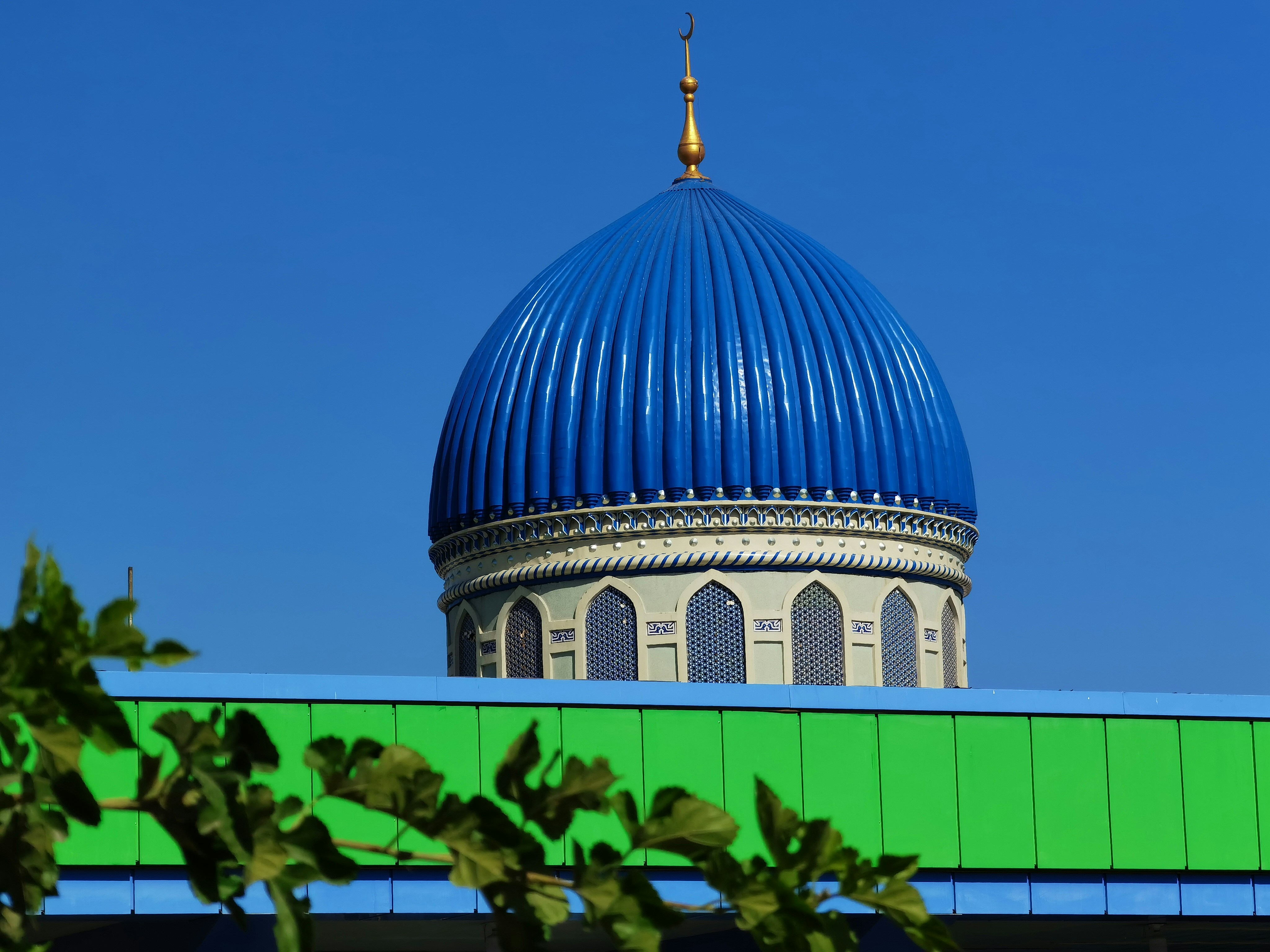 Blue domed structure with ornate windows under a clear sky, partially obscured by green foliage.