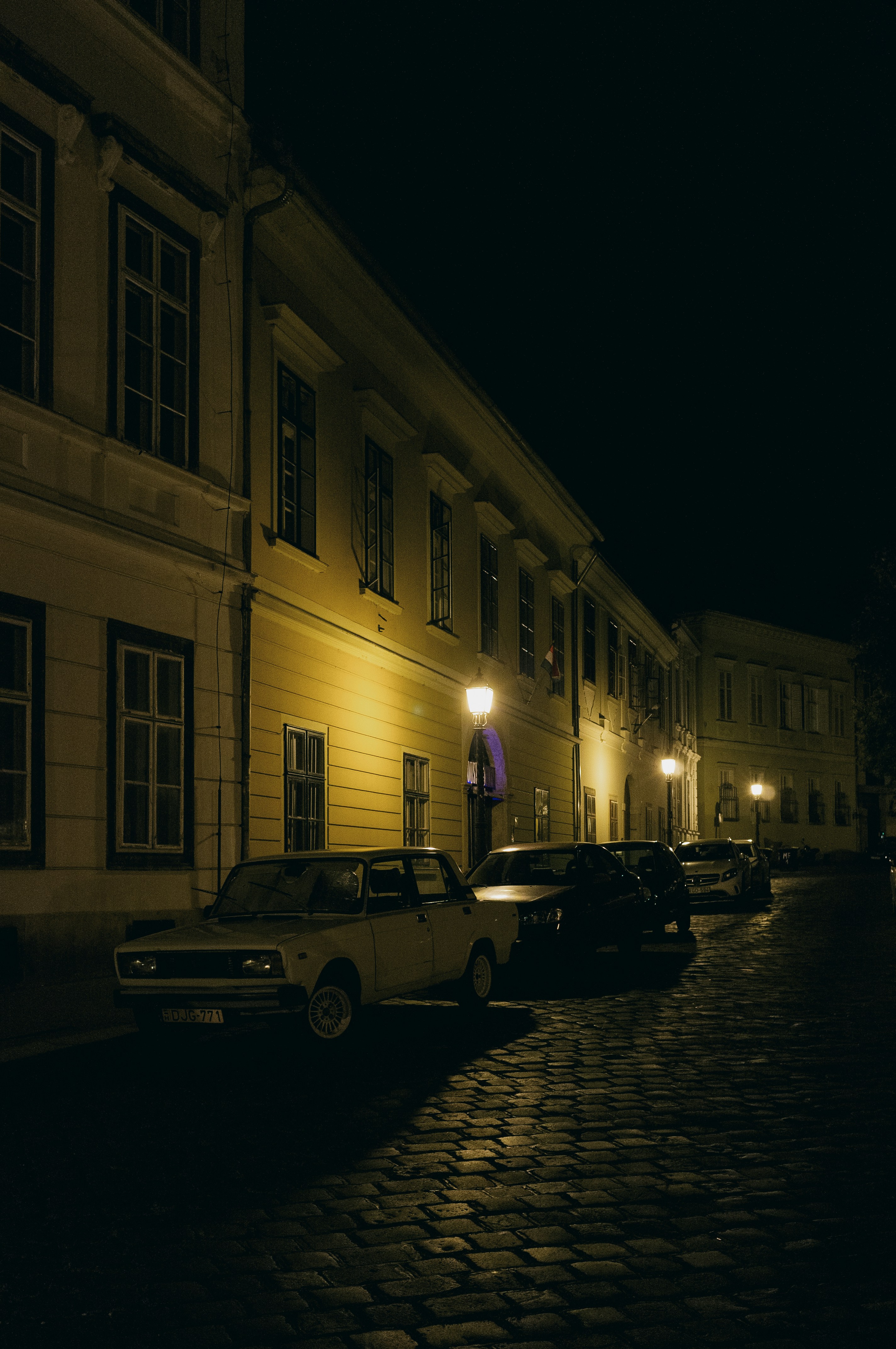 Quiet cobblestone street illuminated by vintage street lamps, showcasing parked cars and historic architecture at night.