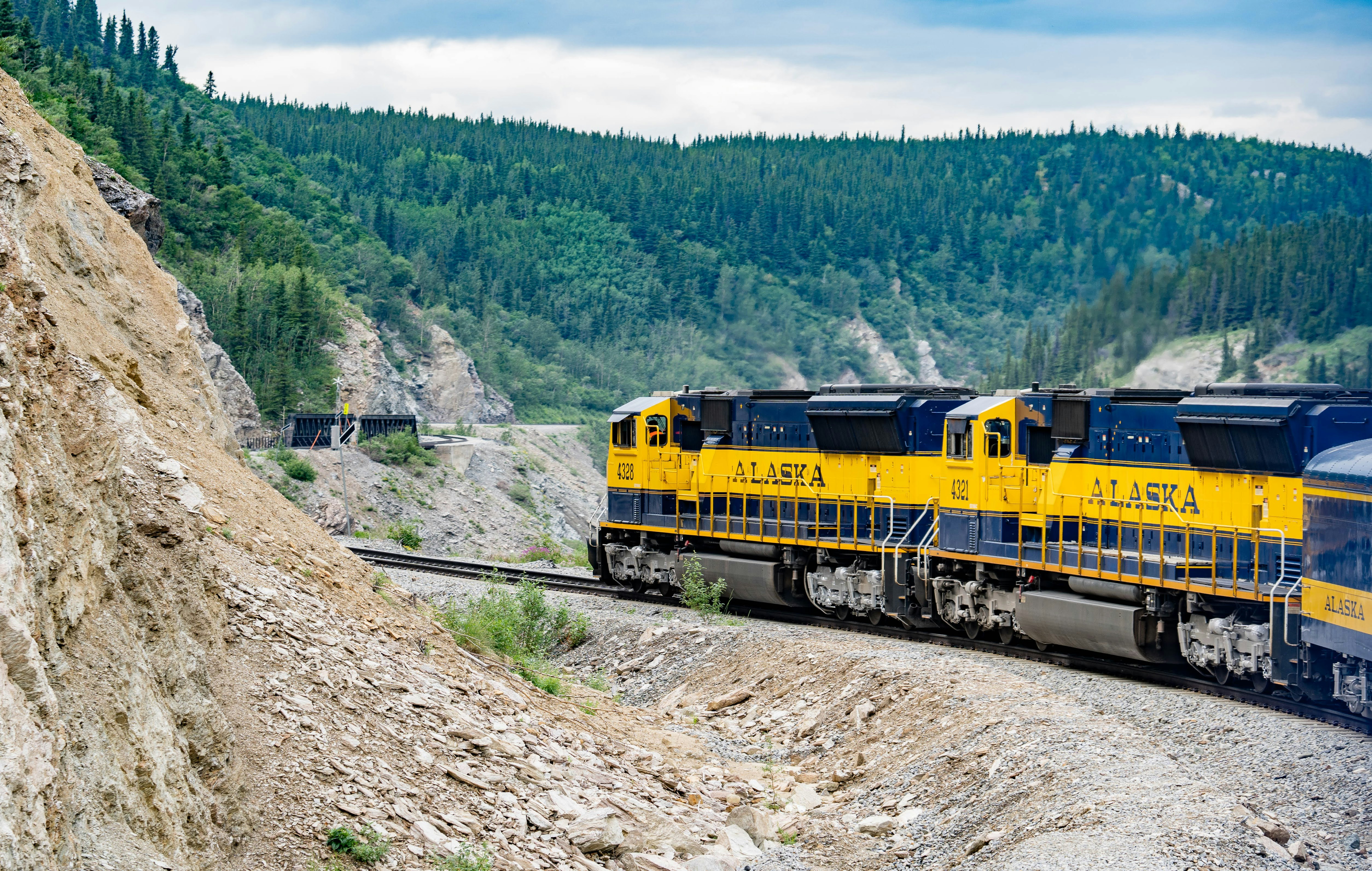Alaska railroad train curves through a lush forested valley beneath a cloudy sky.