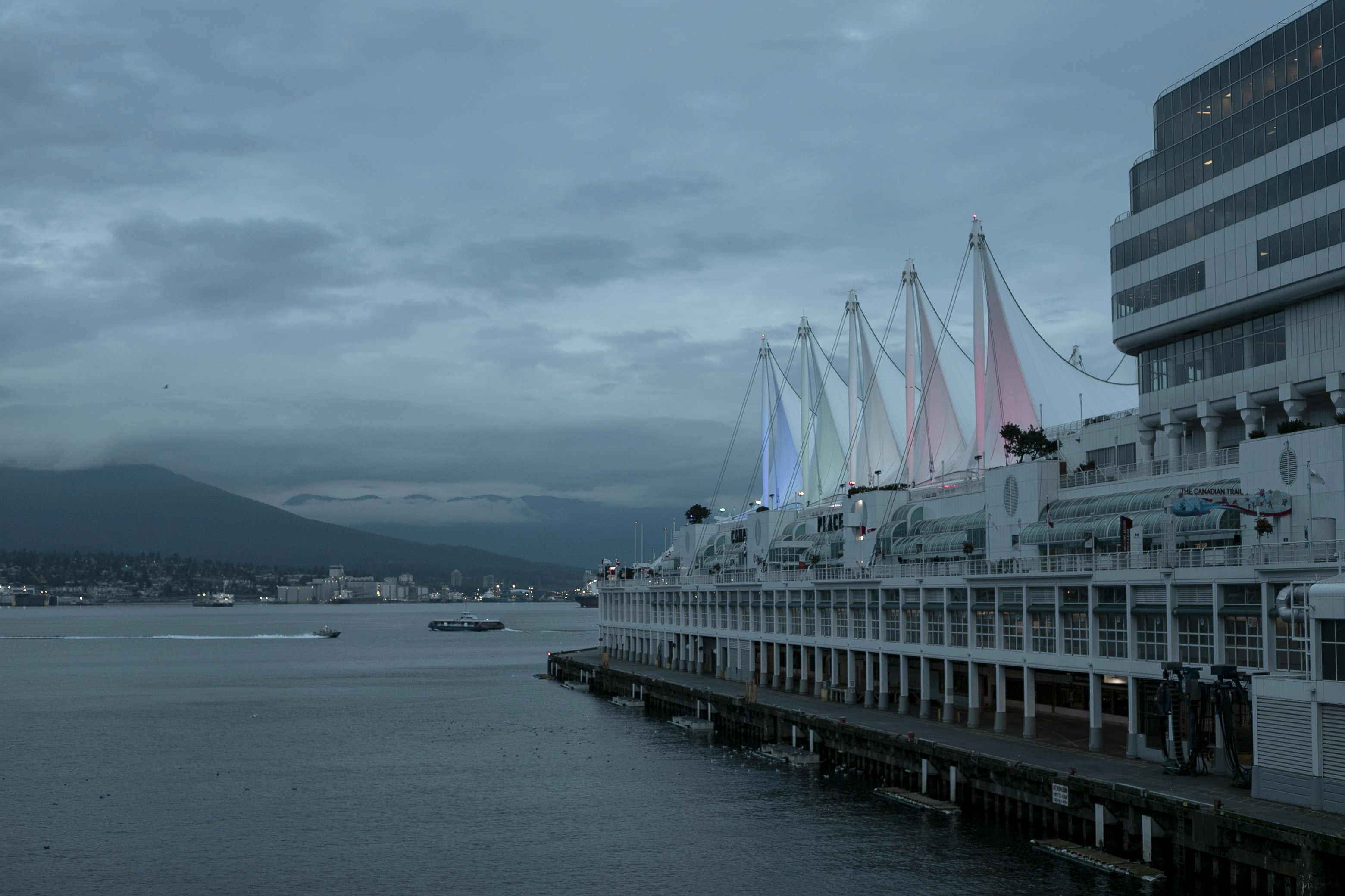 Illuminated harbor structure with colorful sails reflecting in calm waters during twilight. The distant mountains add depth to the tranquil scene.
