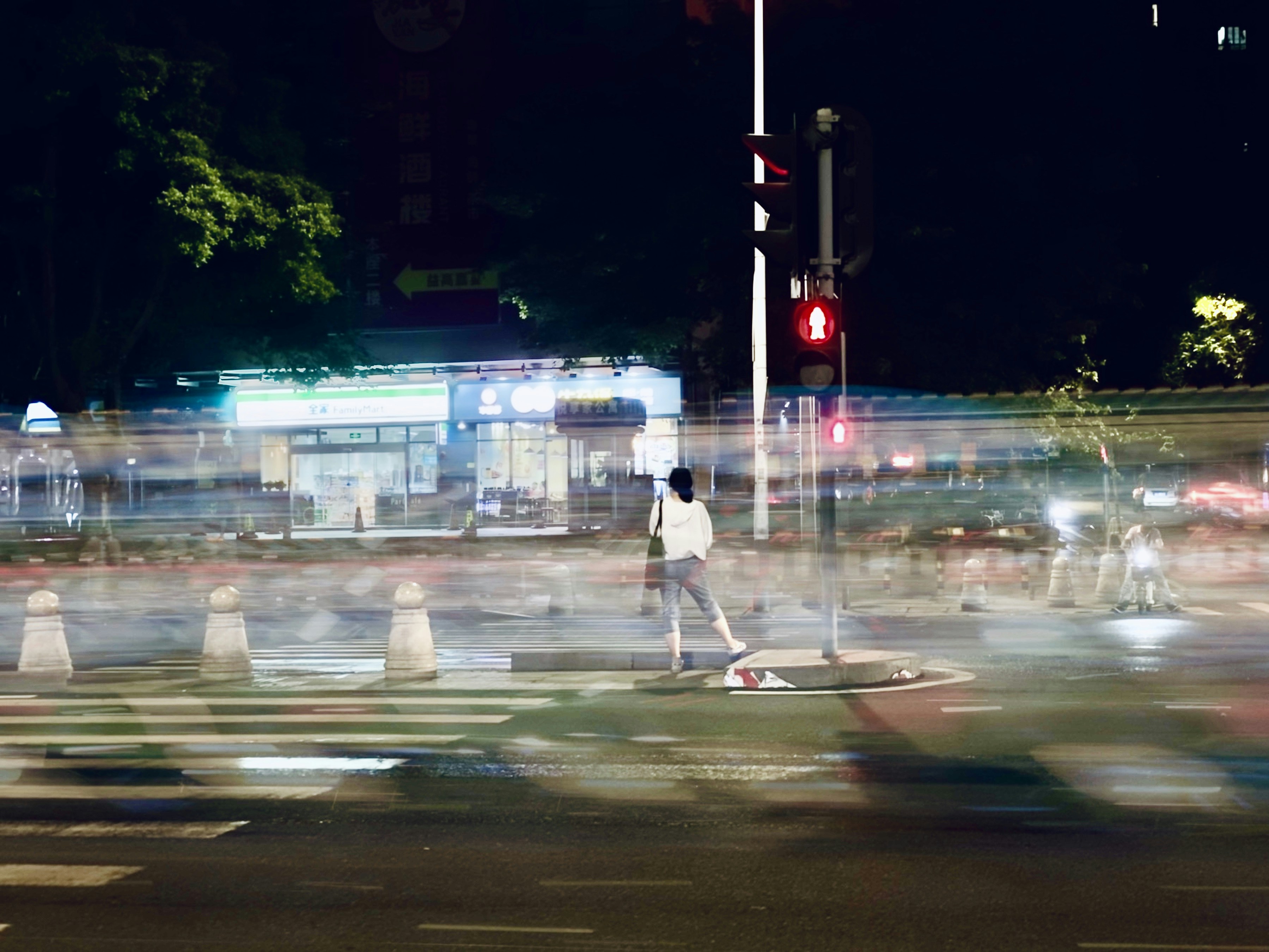 A pedestrian pauses at a crosswalk amidst the swirling lights of passing vehicles at night, creating a dynamic sense of motion.