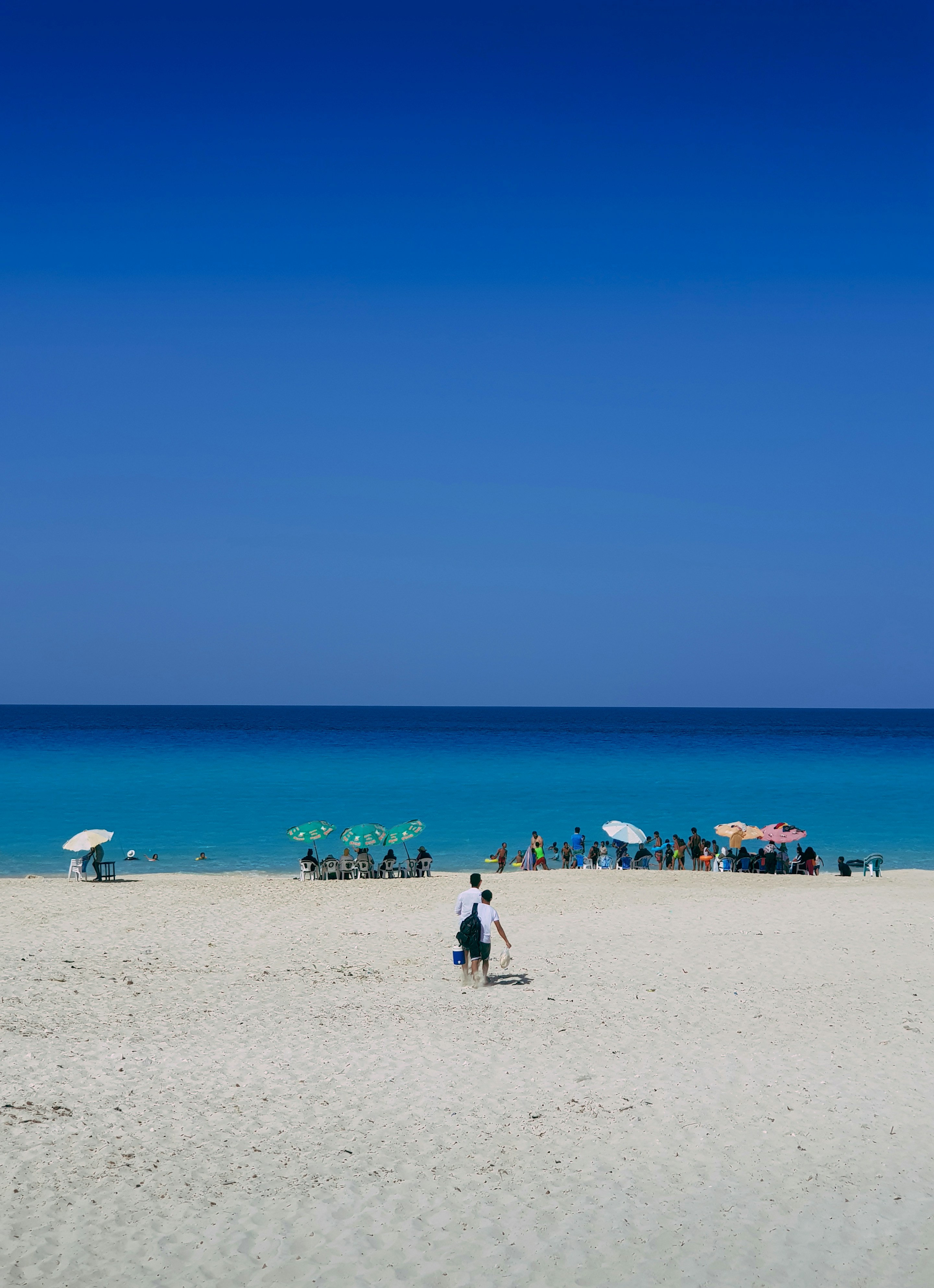 Two figures walk along a sunlit sandy beach toward the turquoise sea under a clear blue sky. Groups of beachgoers and umbrellas line the shore.