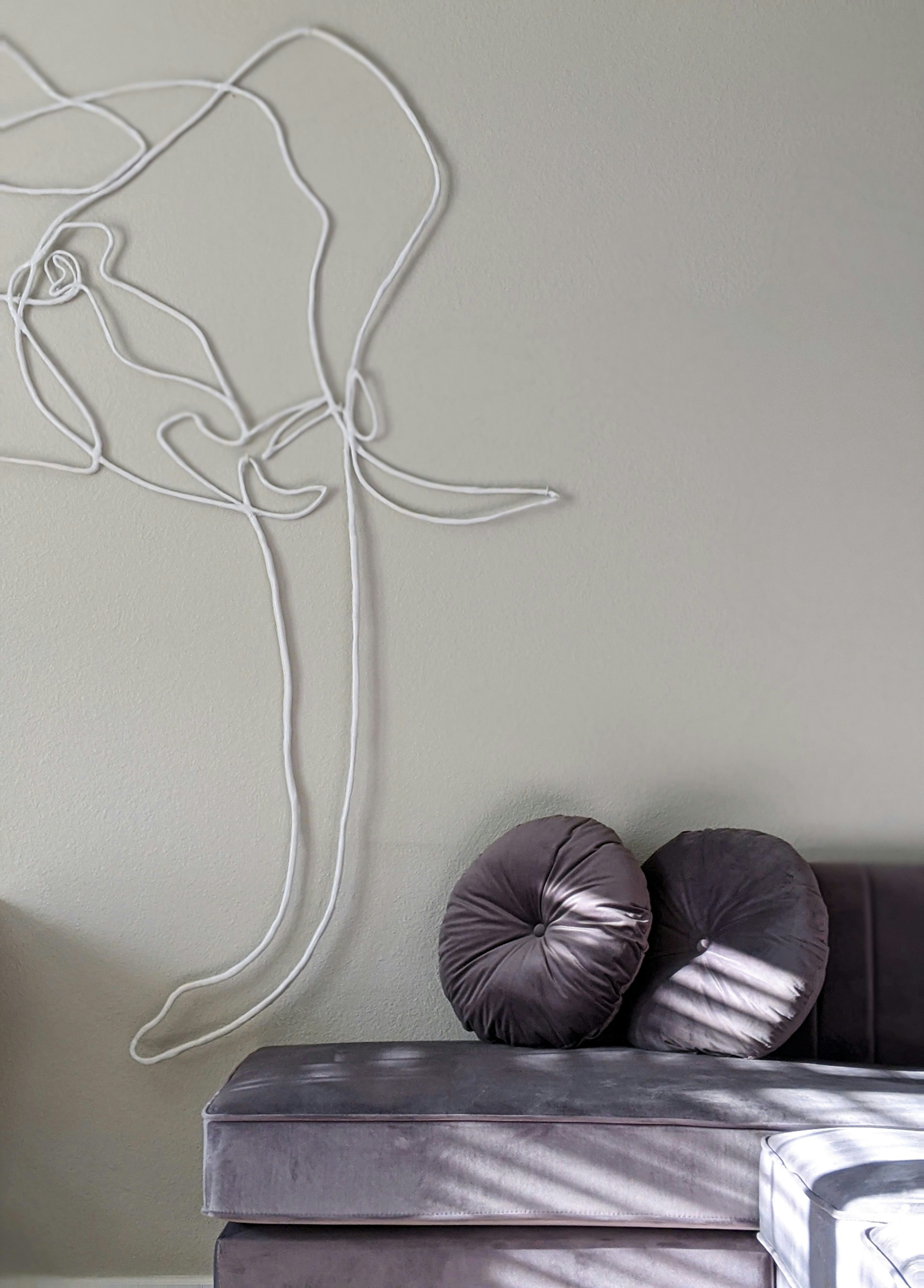 Abstract wall art created with white rope forming a fluid design against a neutral wall, complemented by plush, round cushions on a modern sofa.