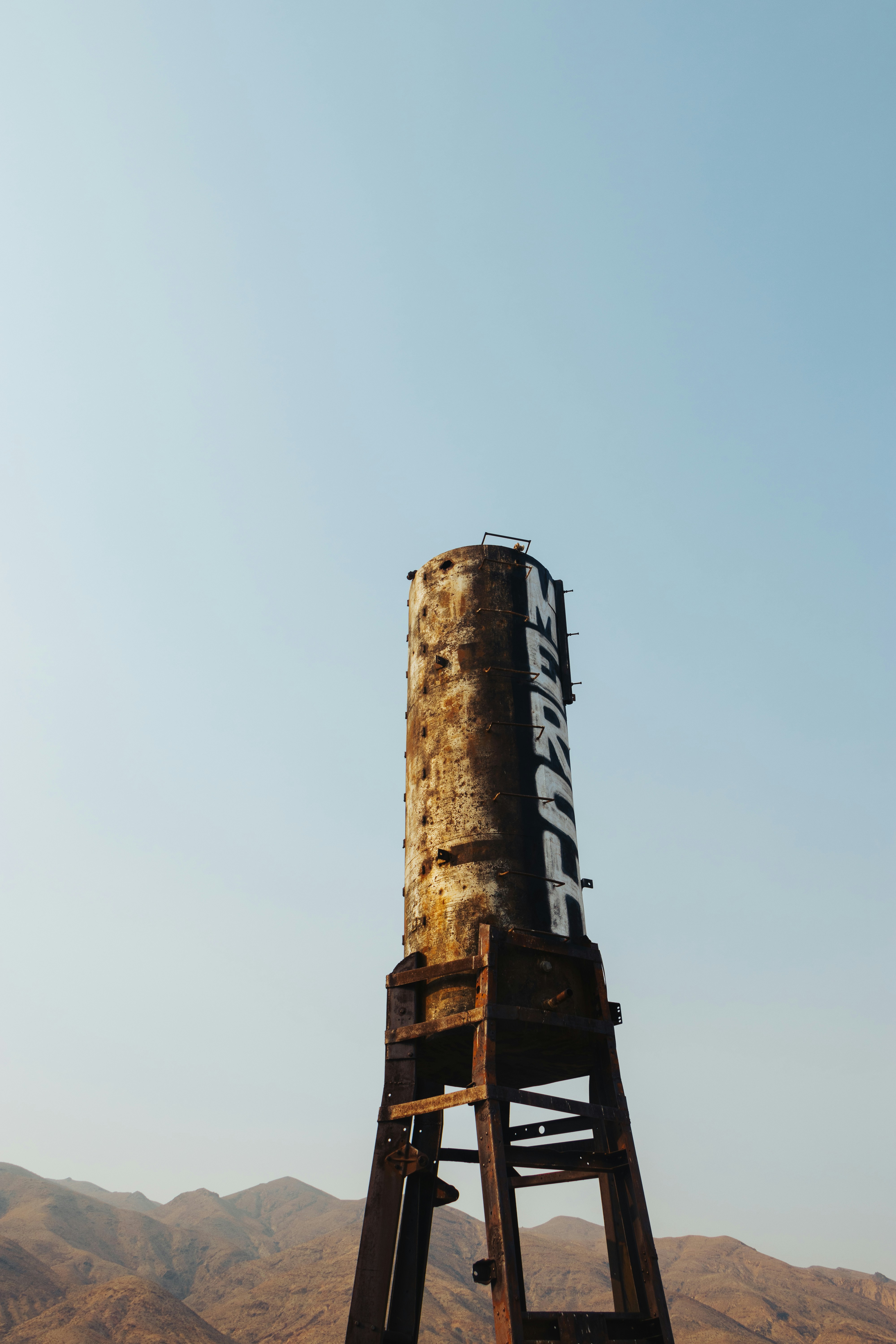 Weathered water tower with faded lettering stands against a clear blue sky, surrounded by rolling hills. The structure symbolizes a bygone era.