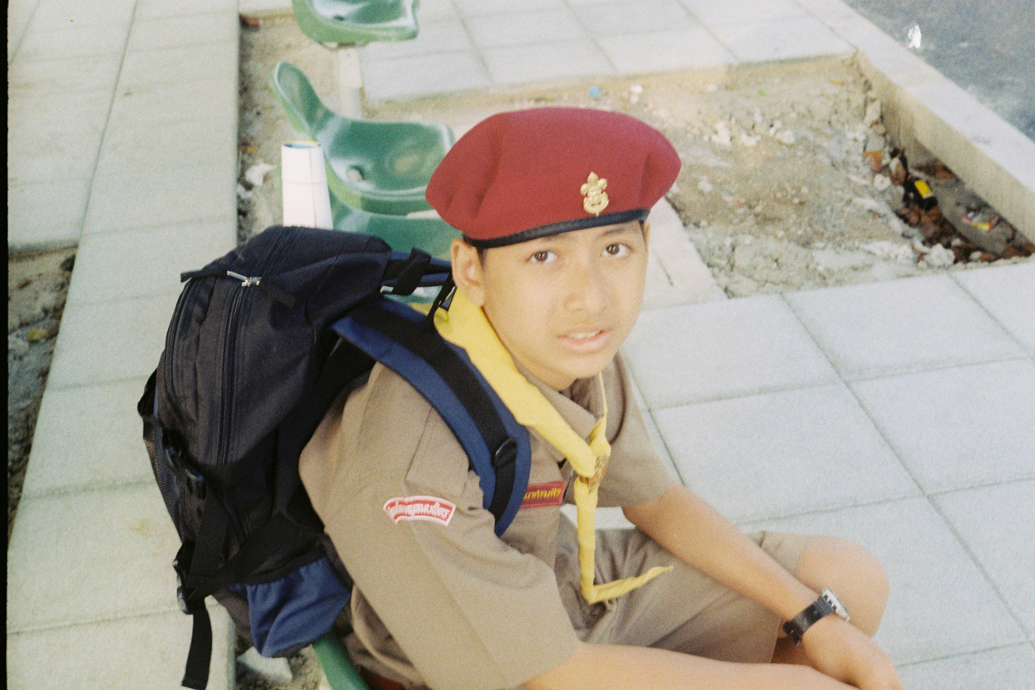 Young scout in uniform with a backpack, seated on a concrete bench, gazing thoughtfully into the distance.