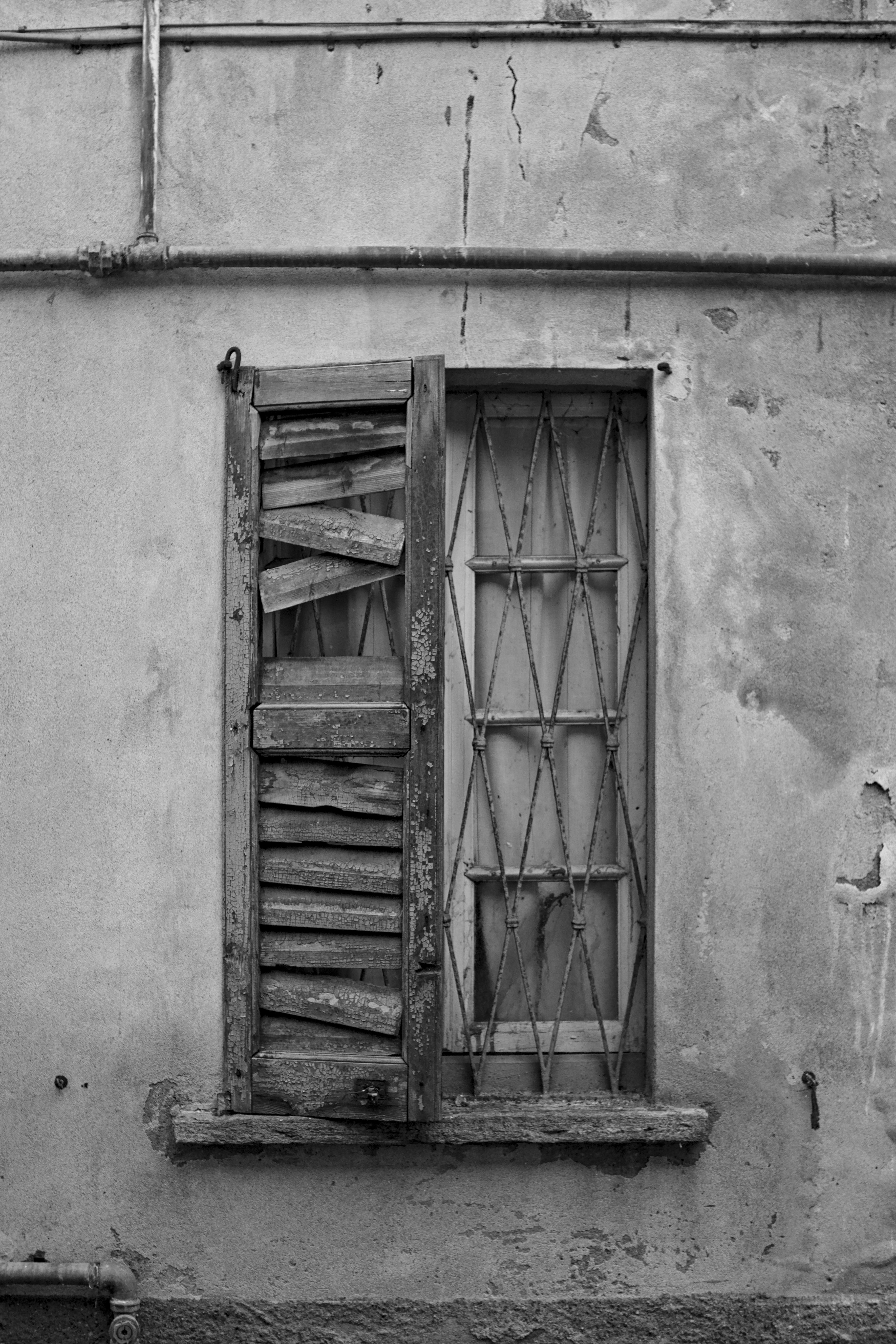 An aged wooden shutter with peeling paint, partially closed against a textured wall, revealing a barred window behind it.