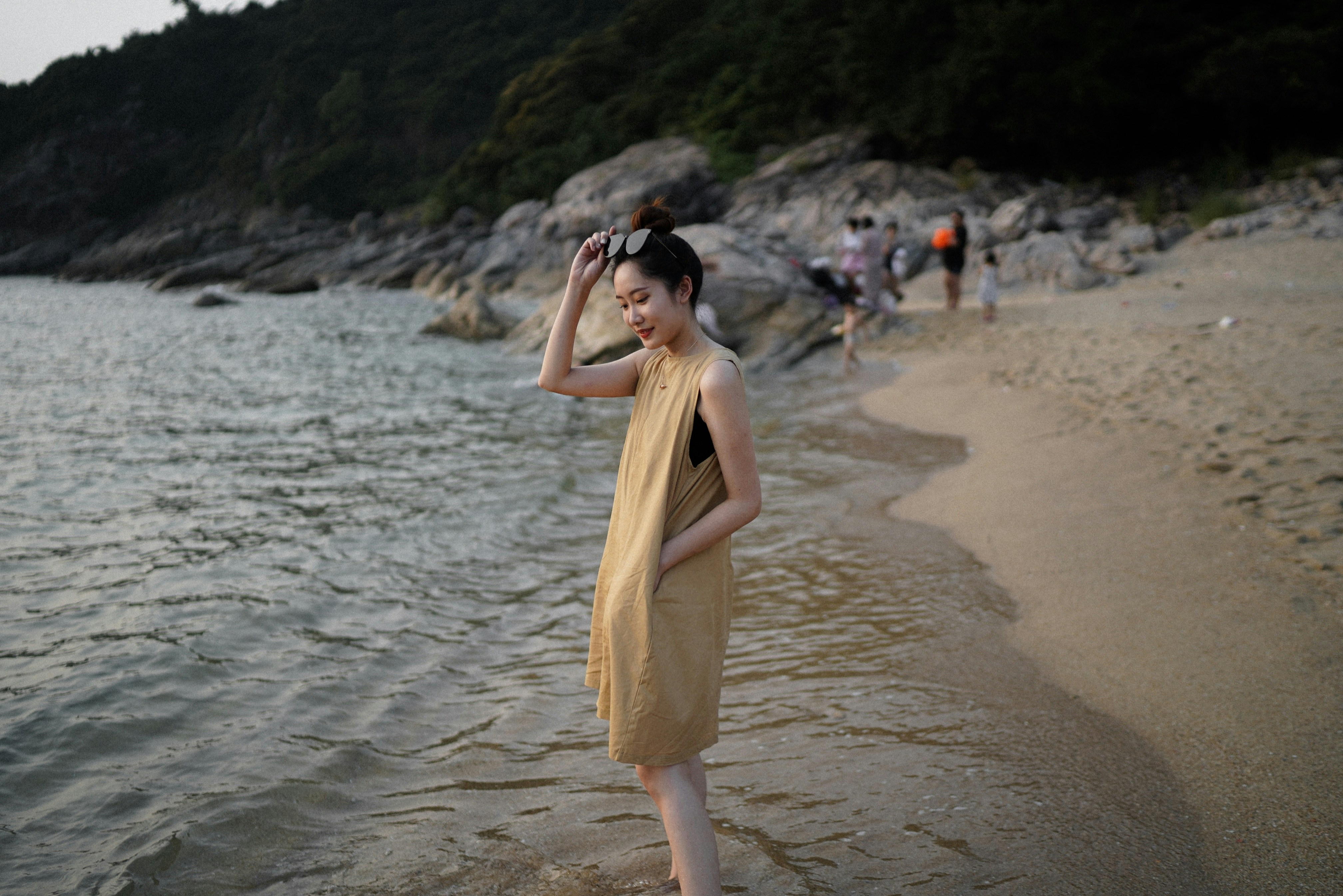 Woman in a golden dress enjoying the gentle waves at the beach, with rocky cliffs in the background. Sunlight casts a warm glow on the scene.