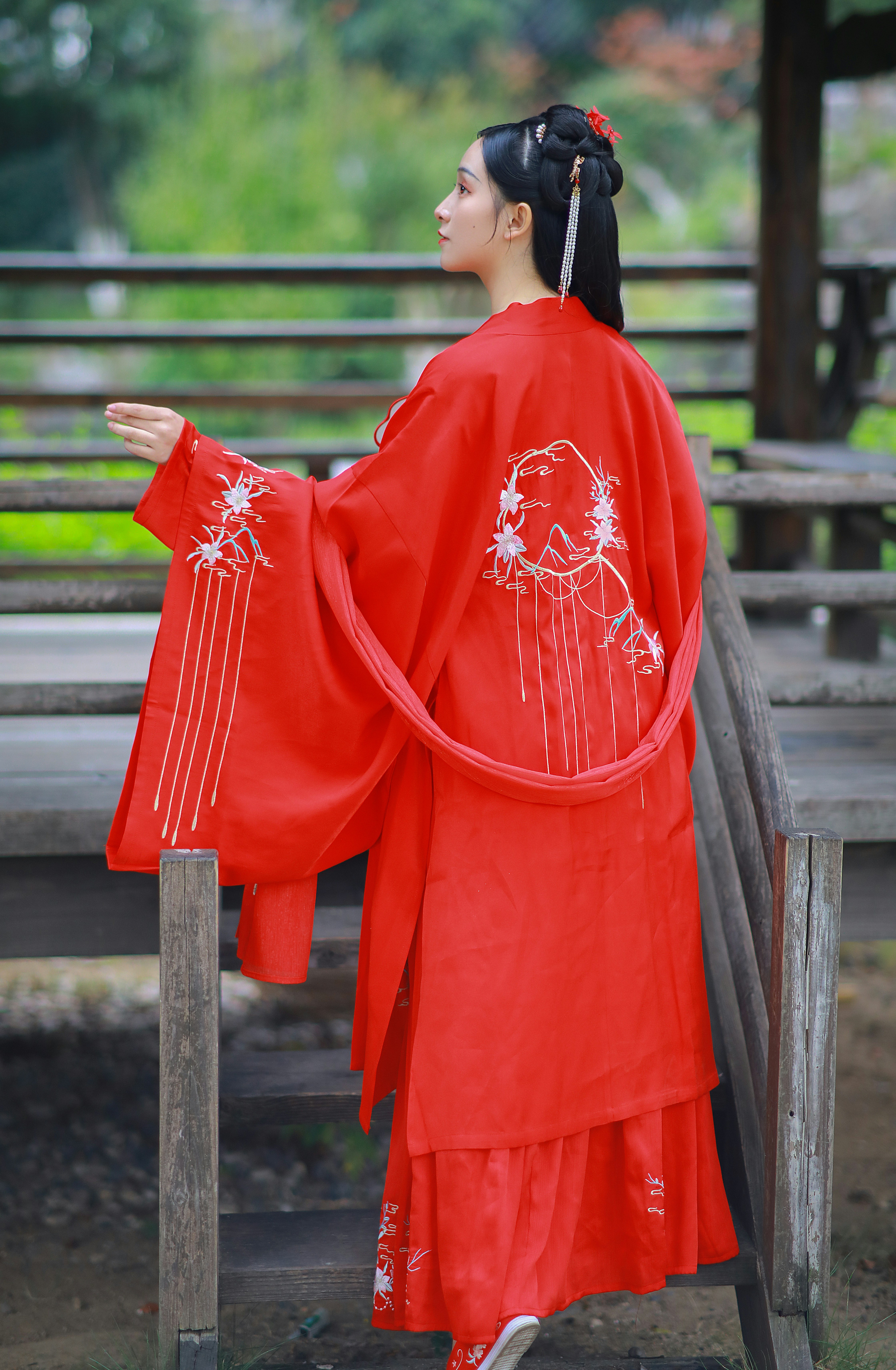 A woman in a flowing red kimono adorned with intricate embroidery stands gracefully on wooden steps, embodying cultural elegance.