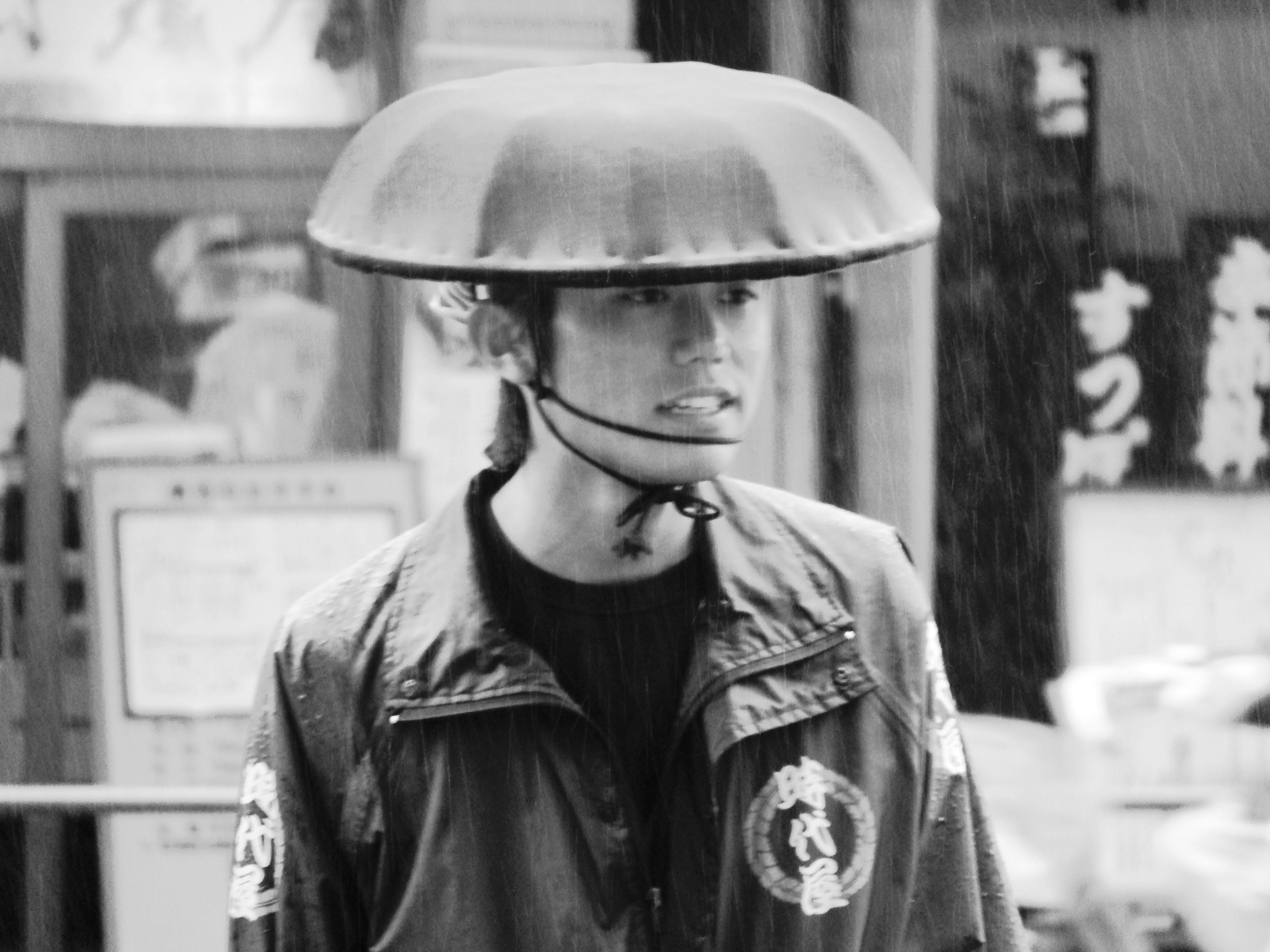 Black-and-white street portrait of a person wearing a broad conical hat in the rain, with an urban backdrop and candid mood.
