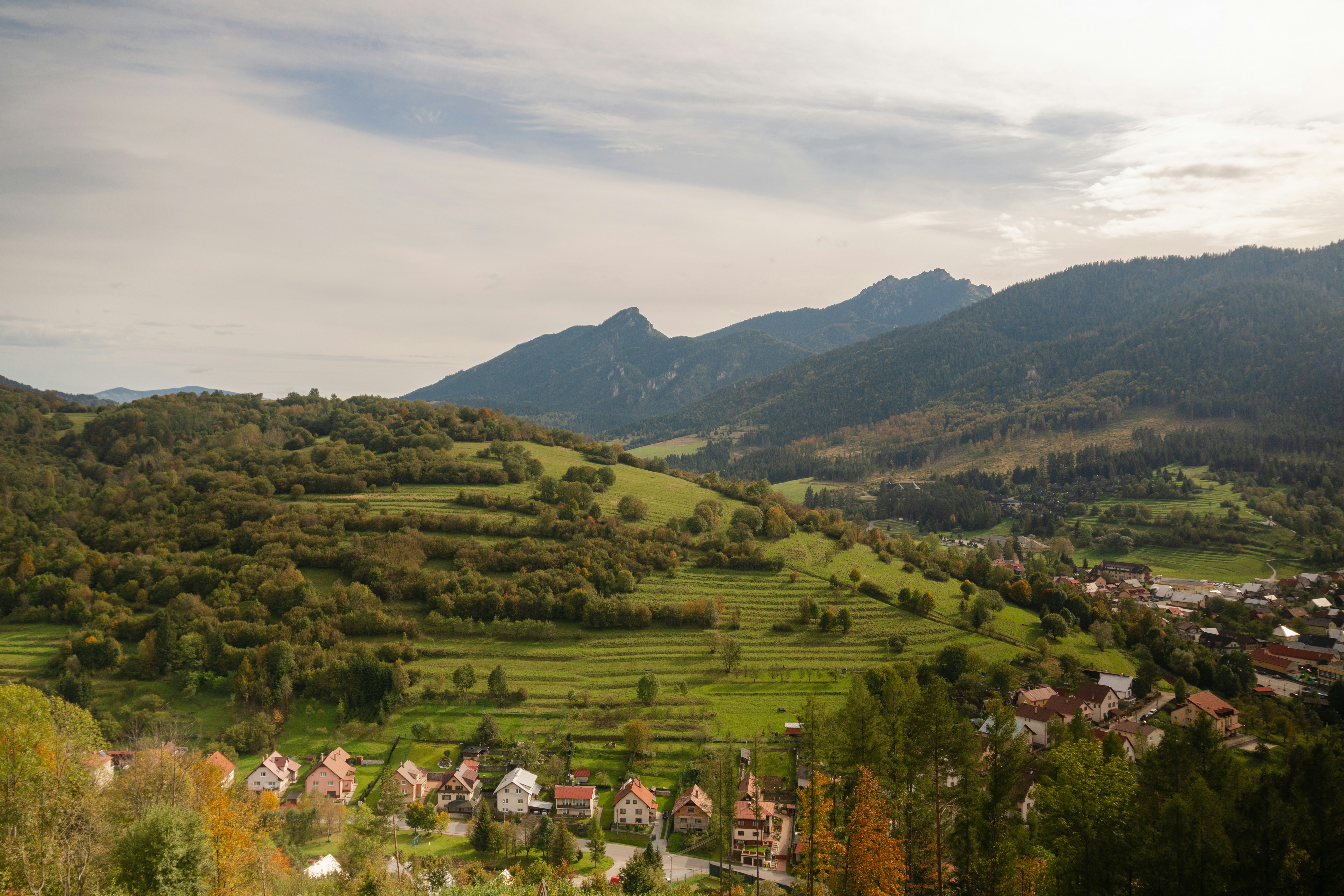 Rolling green fields and clustered houses nestled at the foot of verdant mountains under a cloudy sky.