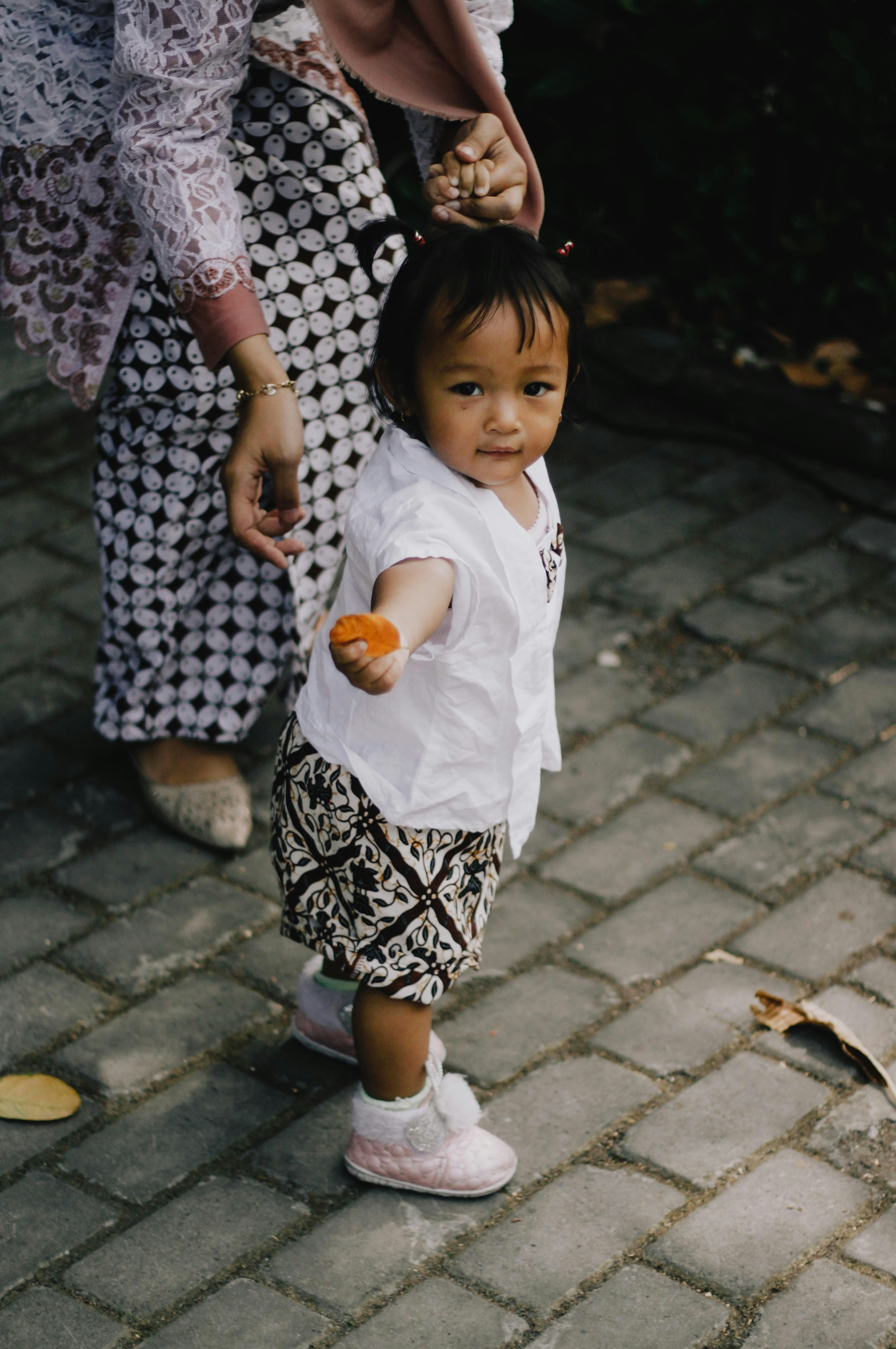 A young child, dressed in a white top and patterned skirt, holds out an orange while standing on a cobblestone path, with a caregiver nearby.