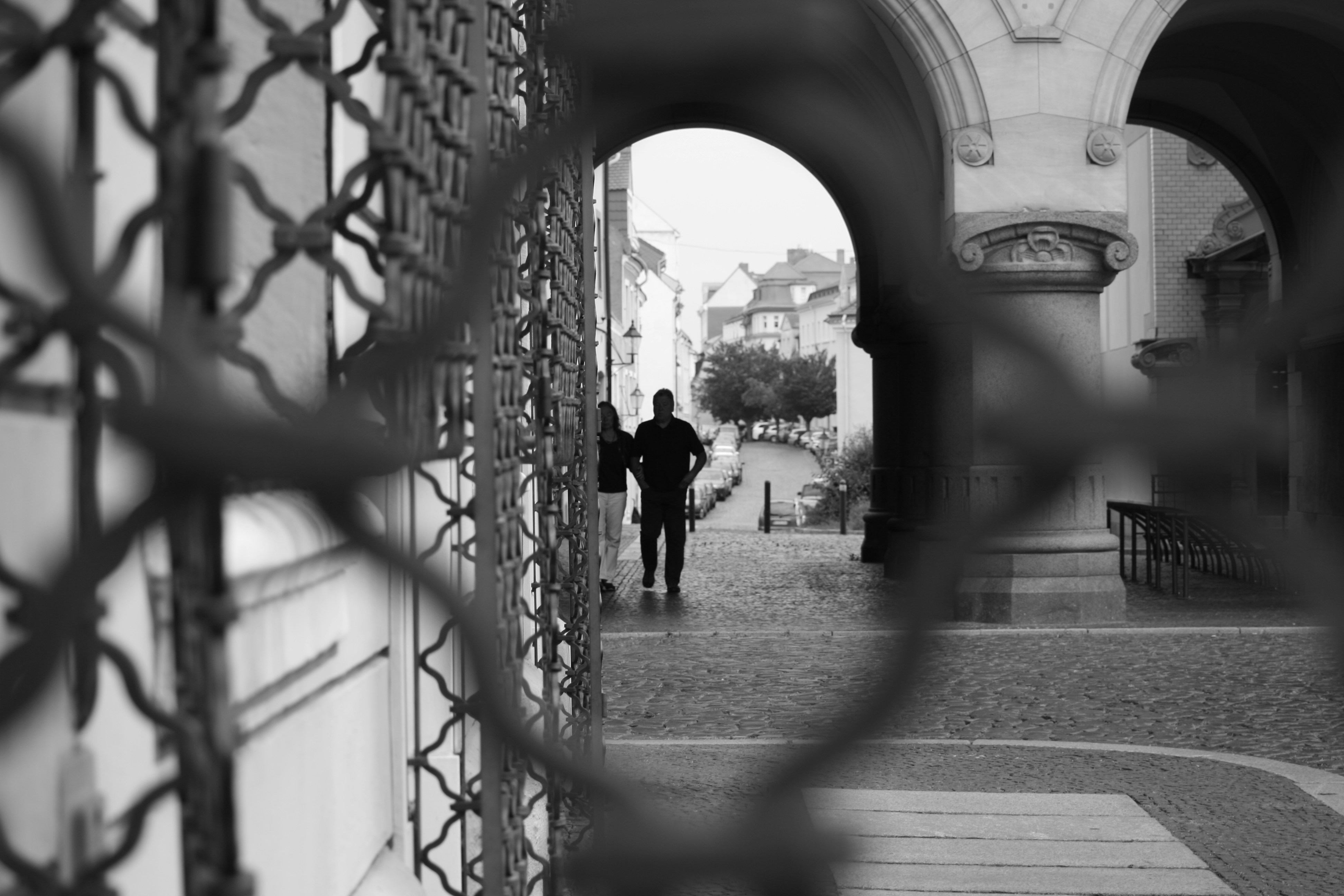 Silhouetted figures walk through an arched gateway framed by intricate wrought iron patterns.
