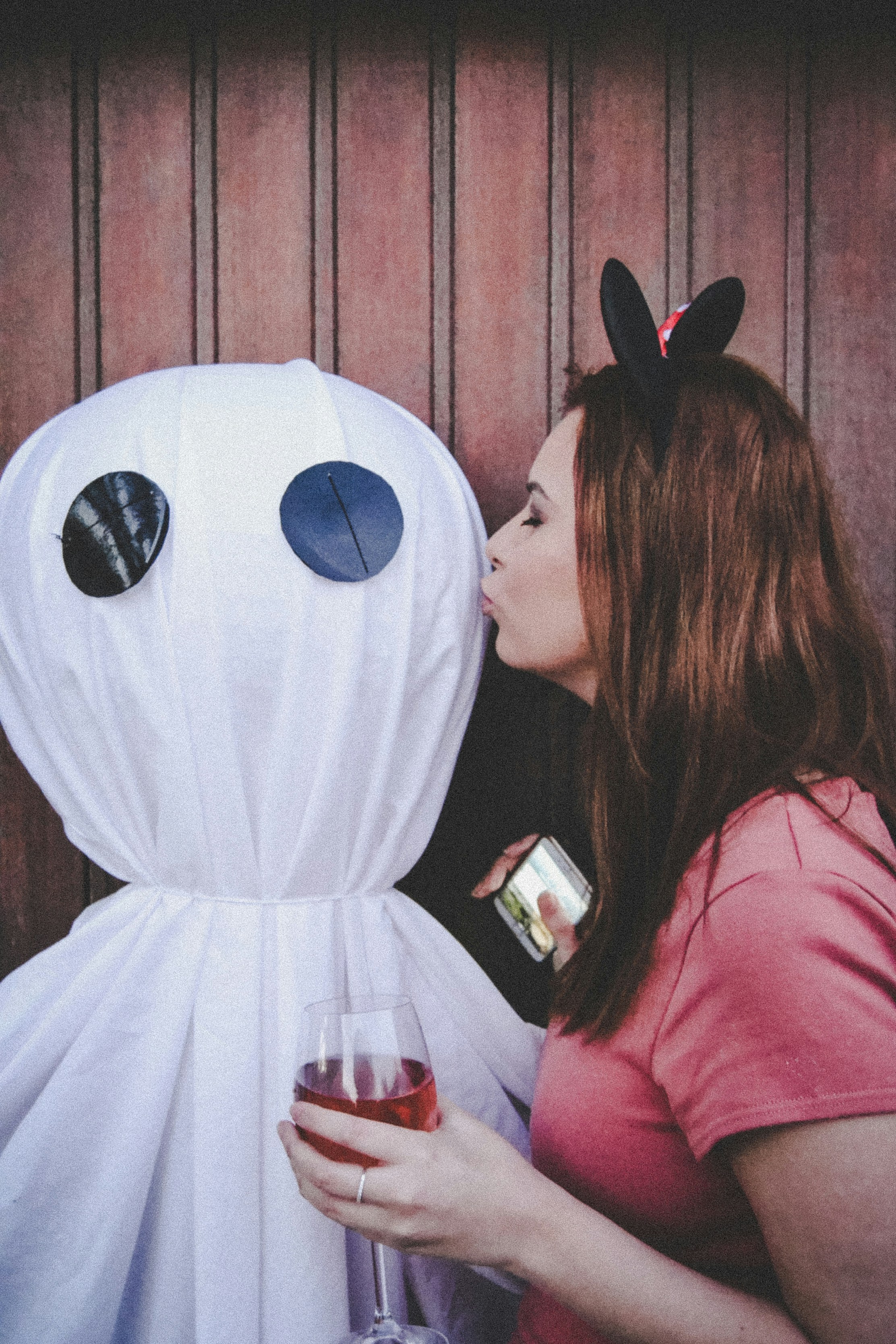 A woman in a pink shirt and black ears leans in to kiss a ghostly figure draped in white, with large black eyes, against a wooden backdrop.