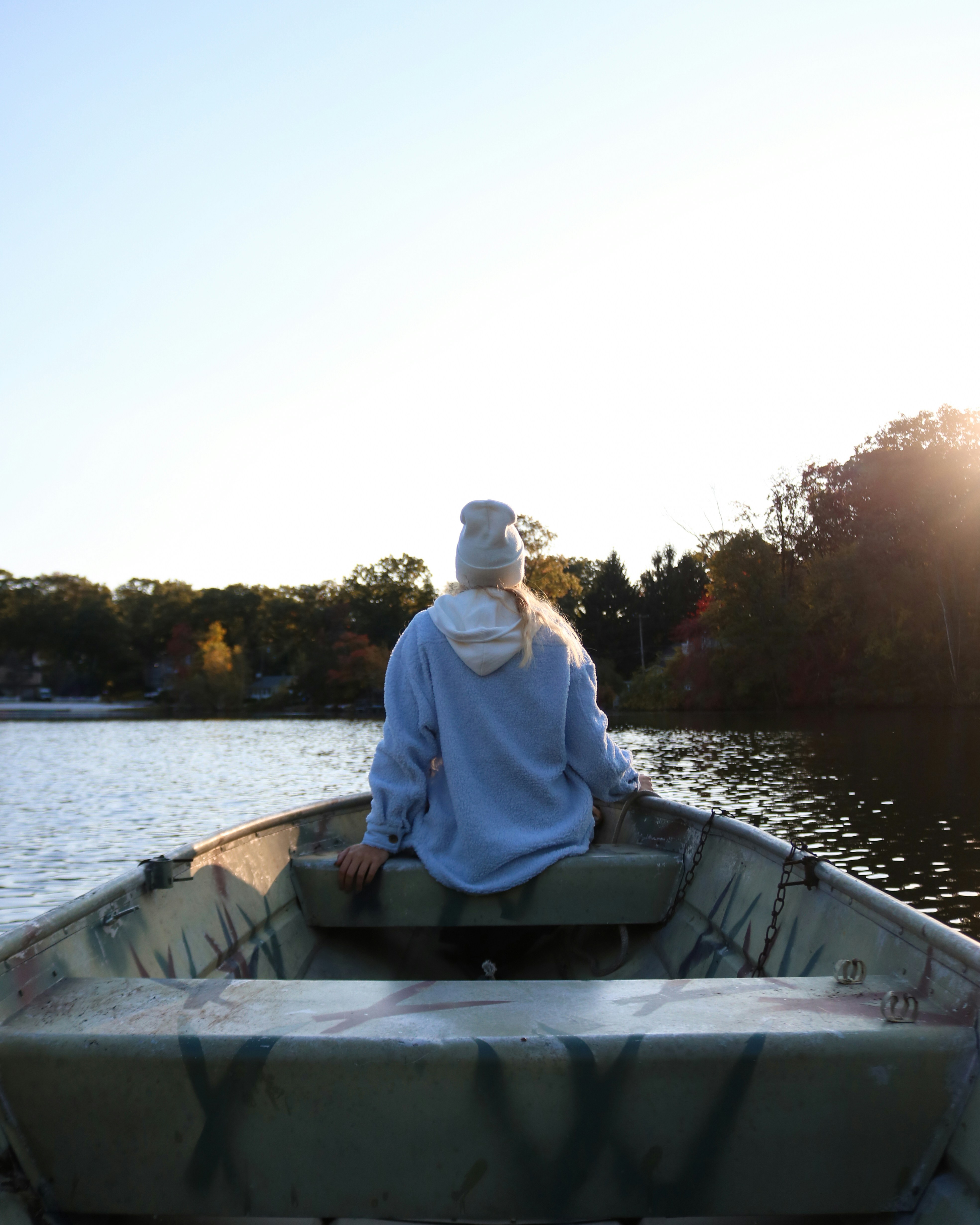 A person sitting in a boat, gazing at the tranquil lake surrounded by autumn foliage. The sun casts a warm glow over the scene.
