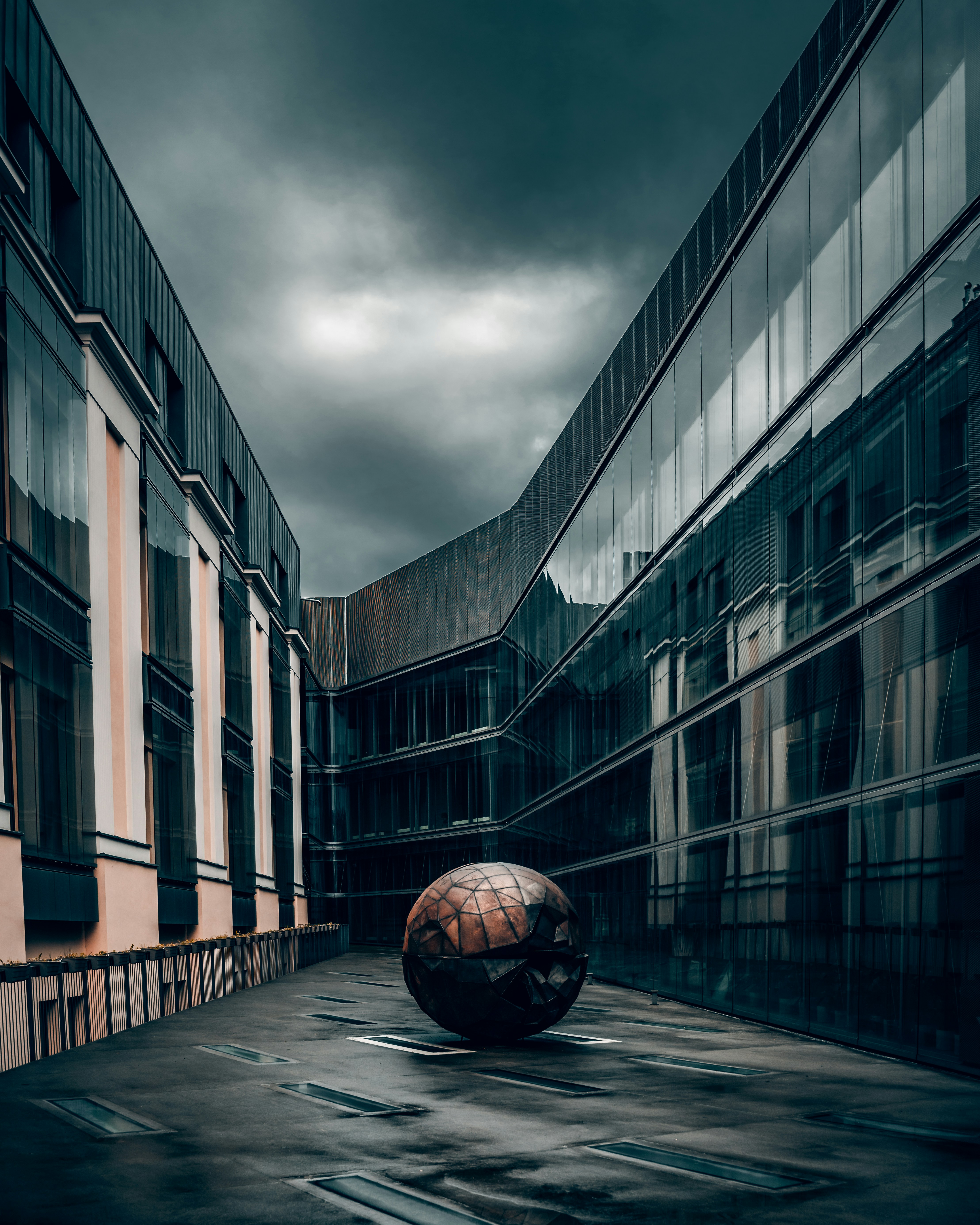 A metallic reflective sphere sculpture sits on a rooftop between glass-walled buildings, capturing the moody sky and surrounding reflections.