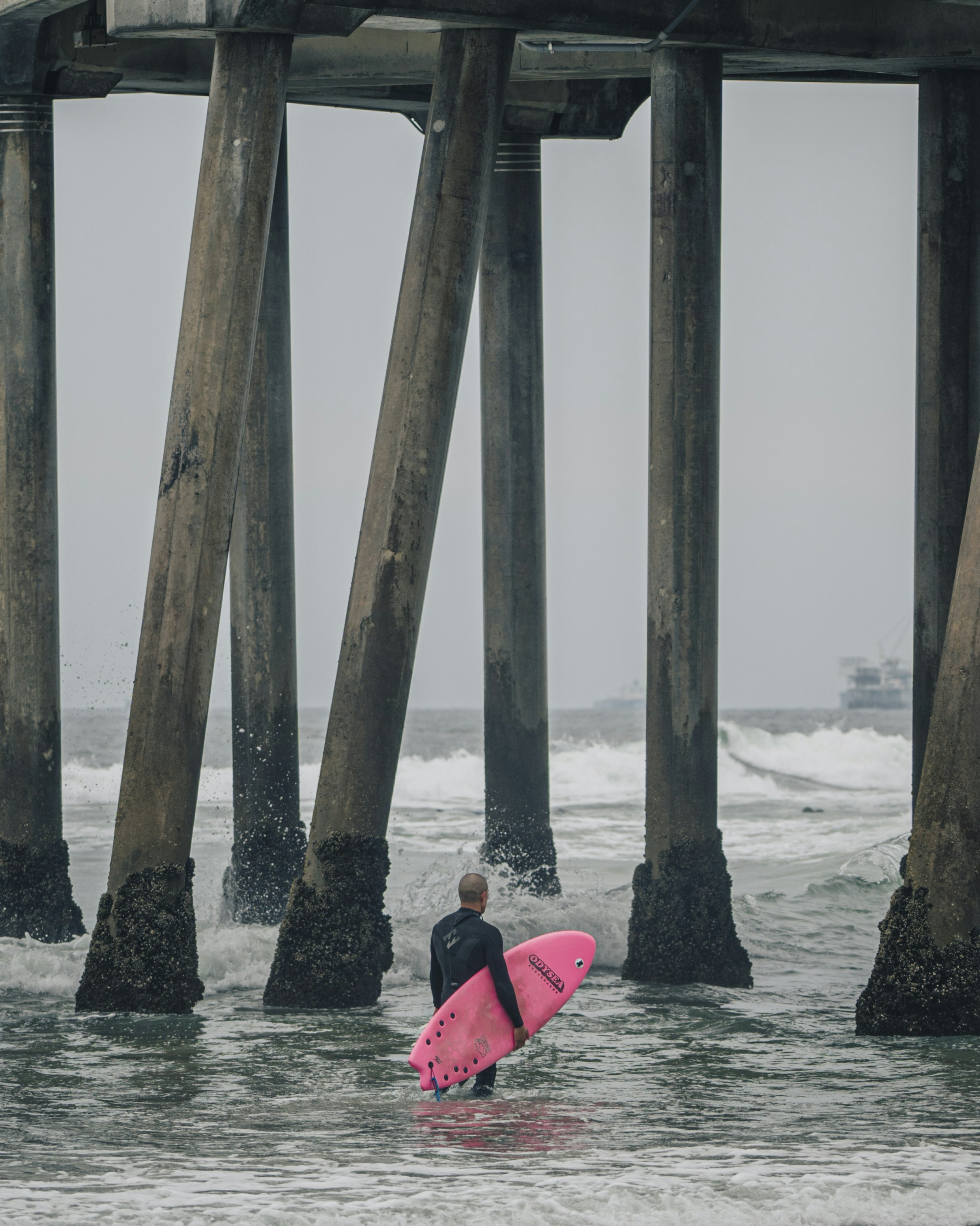 Surfer in a black wetsuit walks toward the ocean, holding a pink surfboard beneath a weathered pier. The scene captures the tranquil yet dynamic relationship between man and nature.