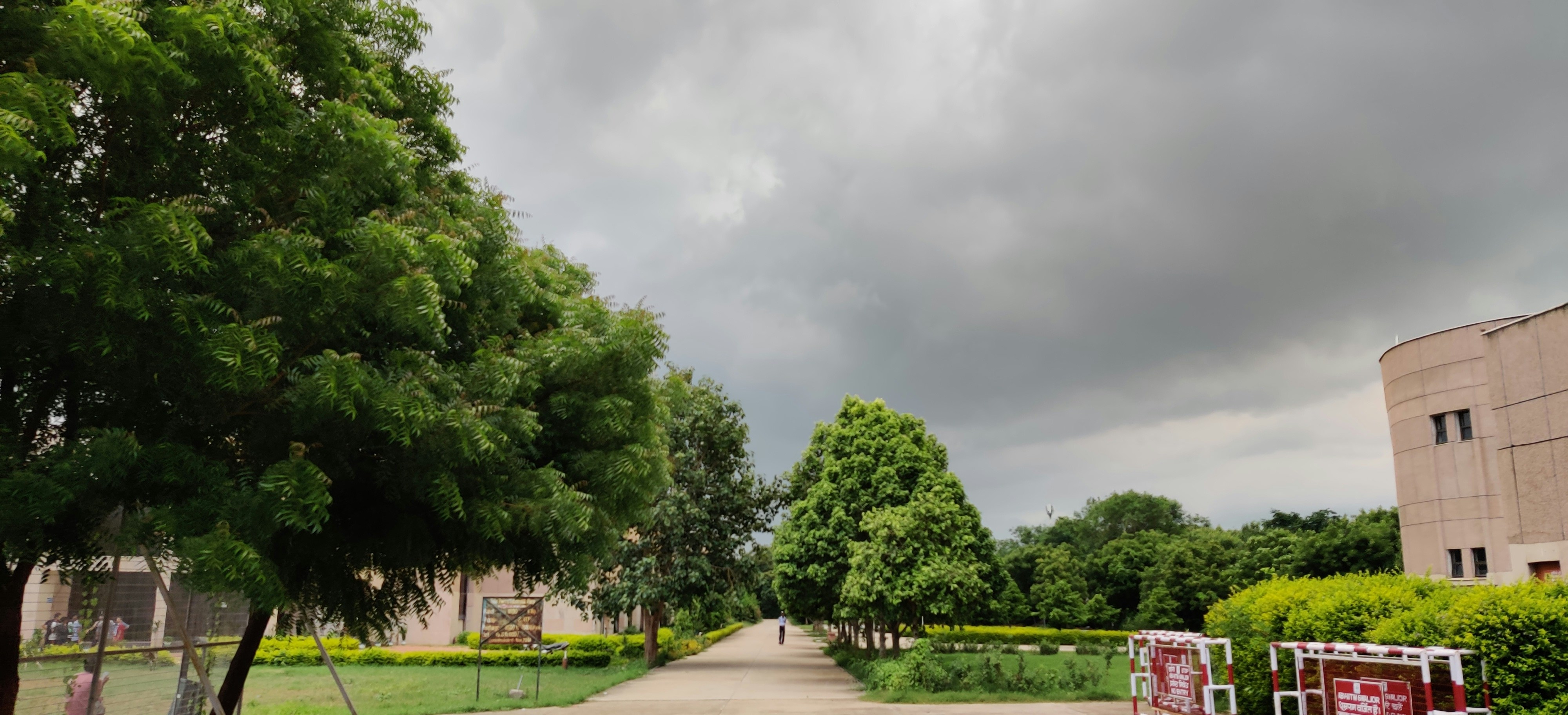 Lush green trees and manicured lawns under a dramatic cloudy sky beside a brick building.