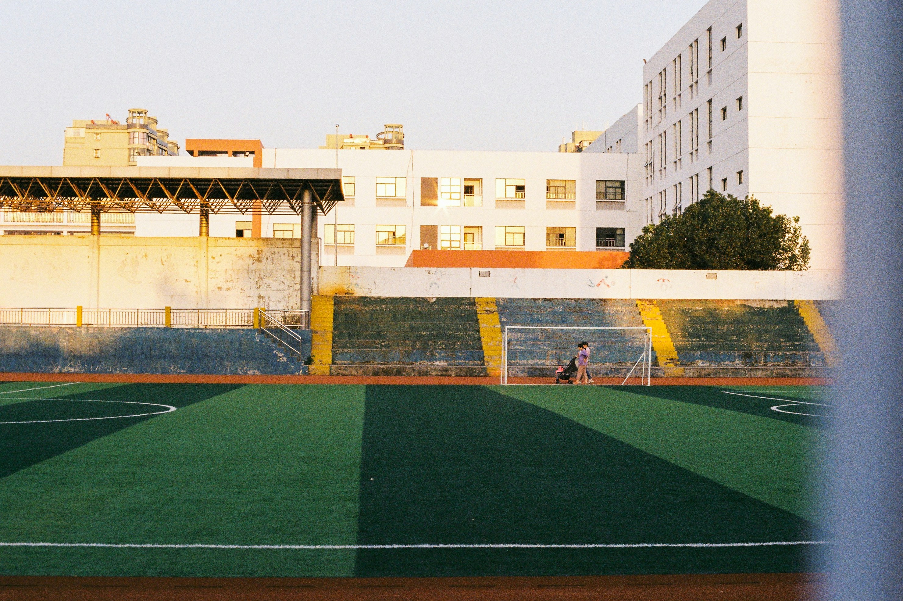 A vibrant soccer field under the warm glow of evening light, featuring empty bleachers and a distant figure preparing for play.