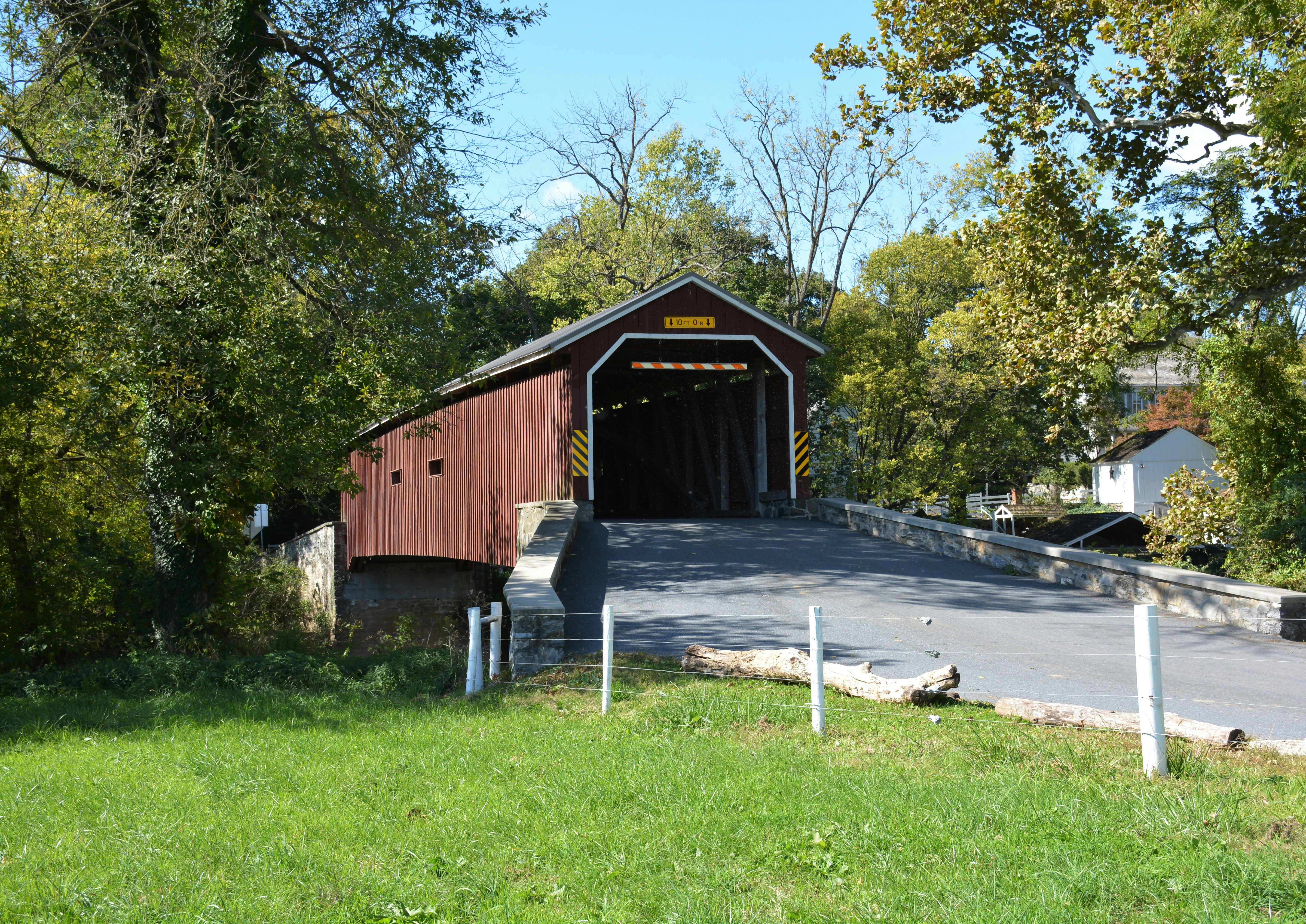 A classic red covered bridge spanning a quiet road, surrounded by lush greenery and trees under a clear blue sky.