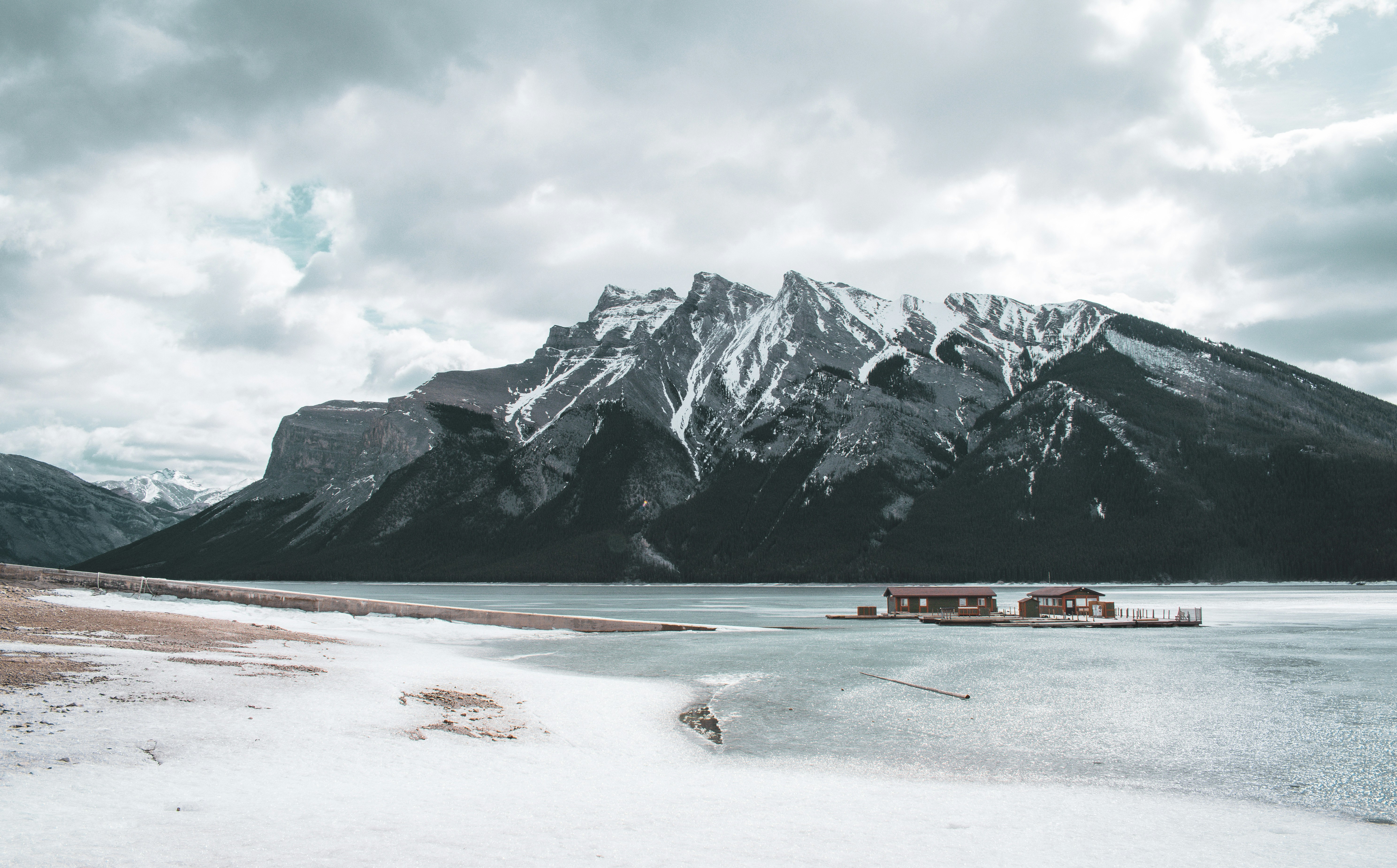 Snow-covered mountains loom over a tranquil lake with a solitary cabin, evoking a sense of solitude and peace.