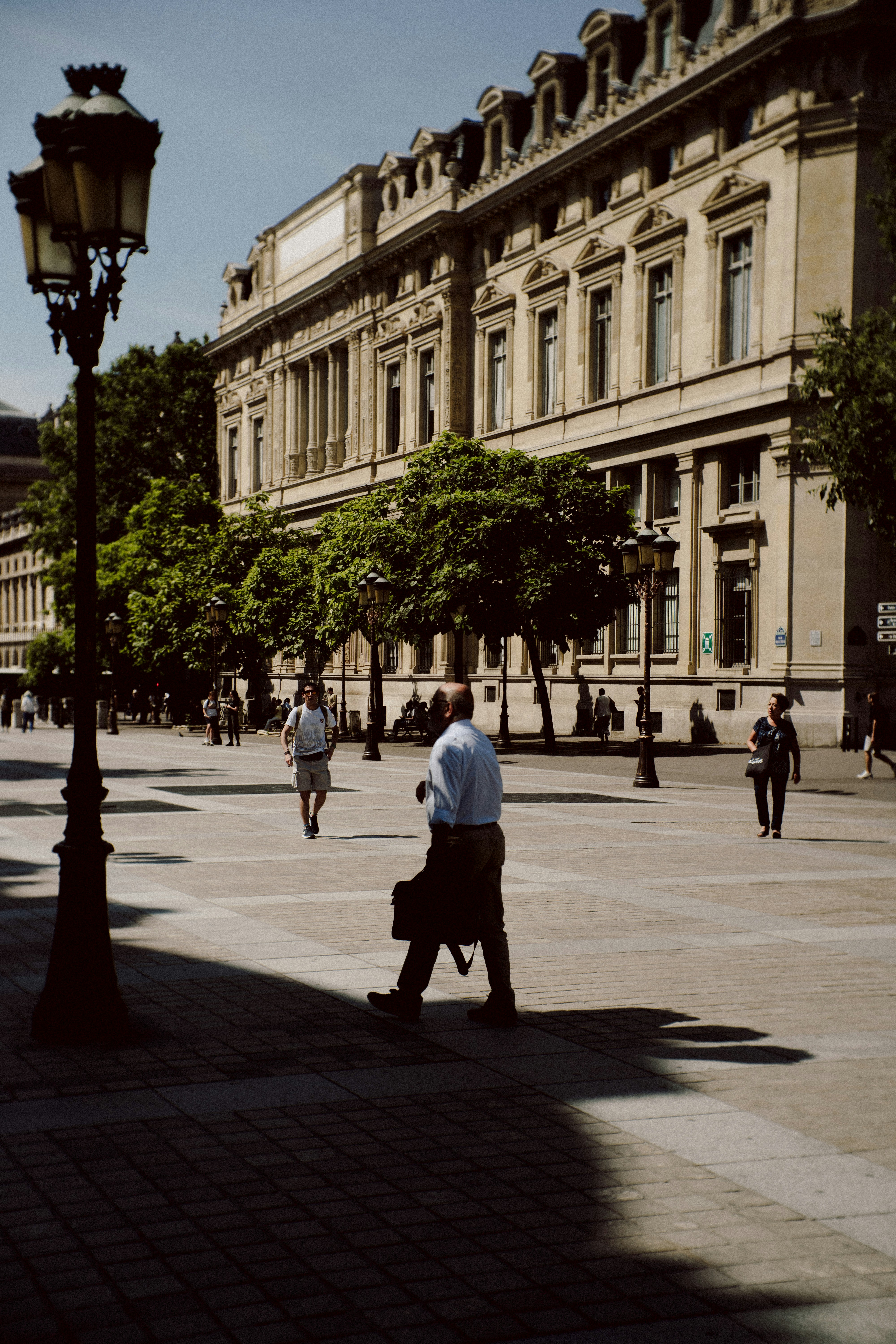 A man walks with a briefcase on a sunlit plaza, framed by lush trees and historic buildings. The scene captures the essence of city life.