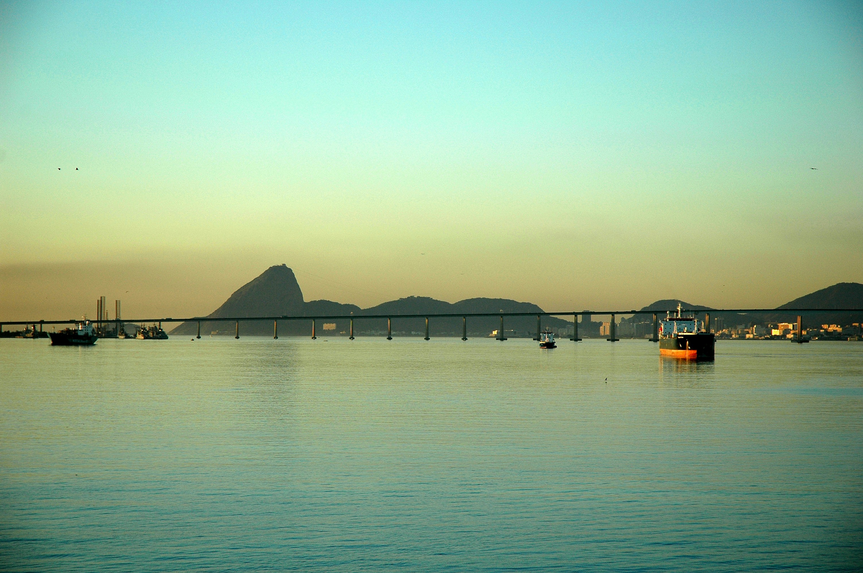 A tranquil view of a bridge and boats on calm waters at dusk, with a distant mountain silhouette. The soft hues of the sky reflect on the water's surface.