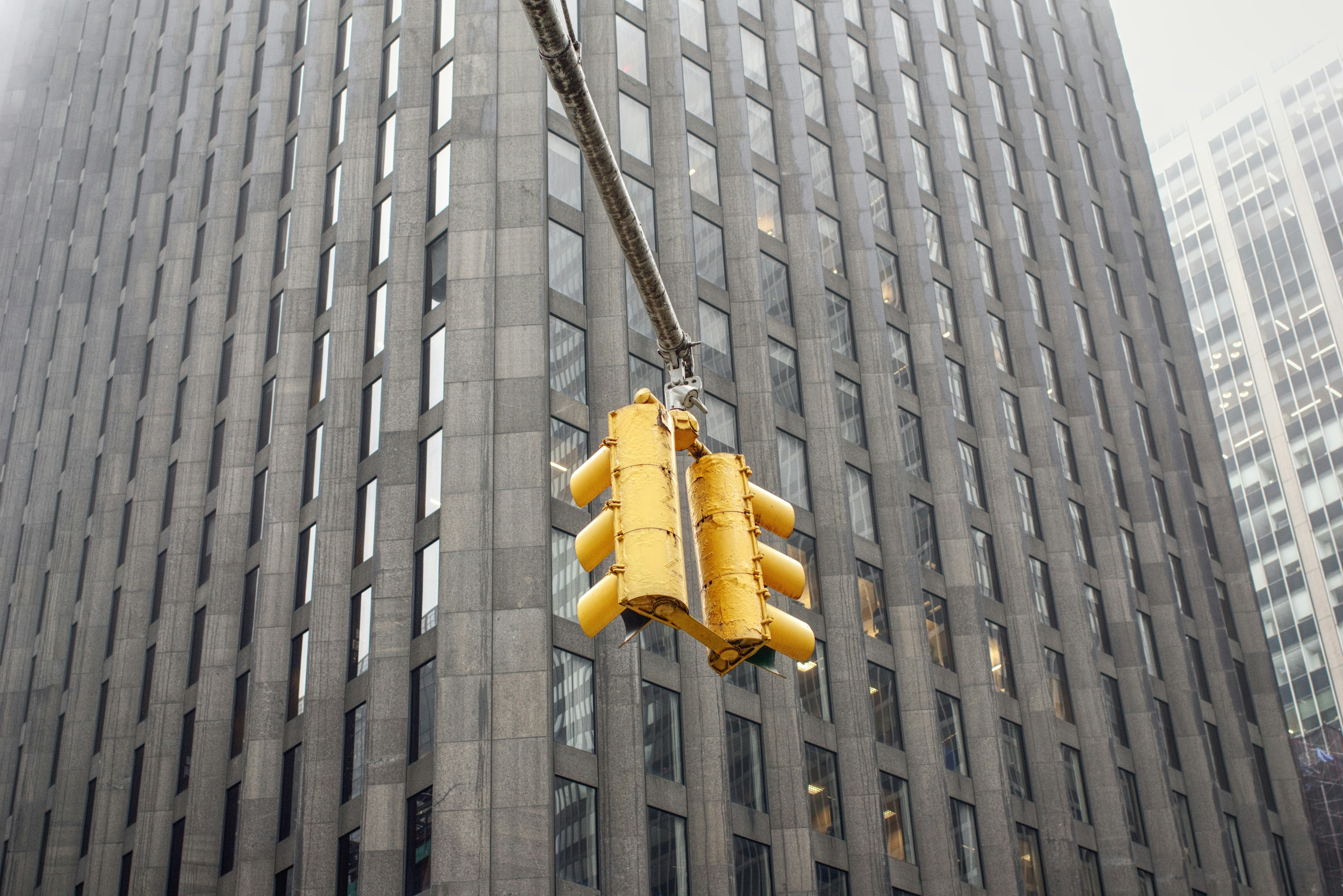 Yellow traffic light suspended against a backdrop of towering gray skyscrapers in a foggy urban setting.