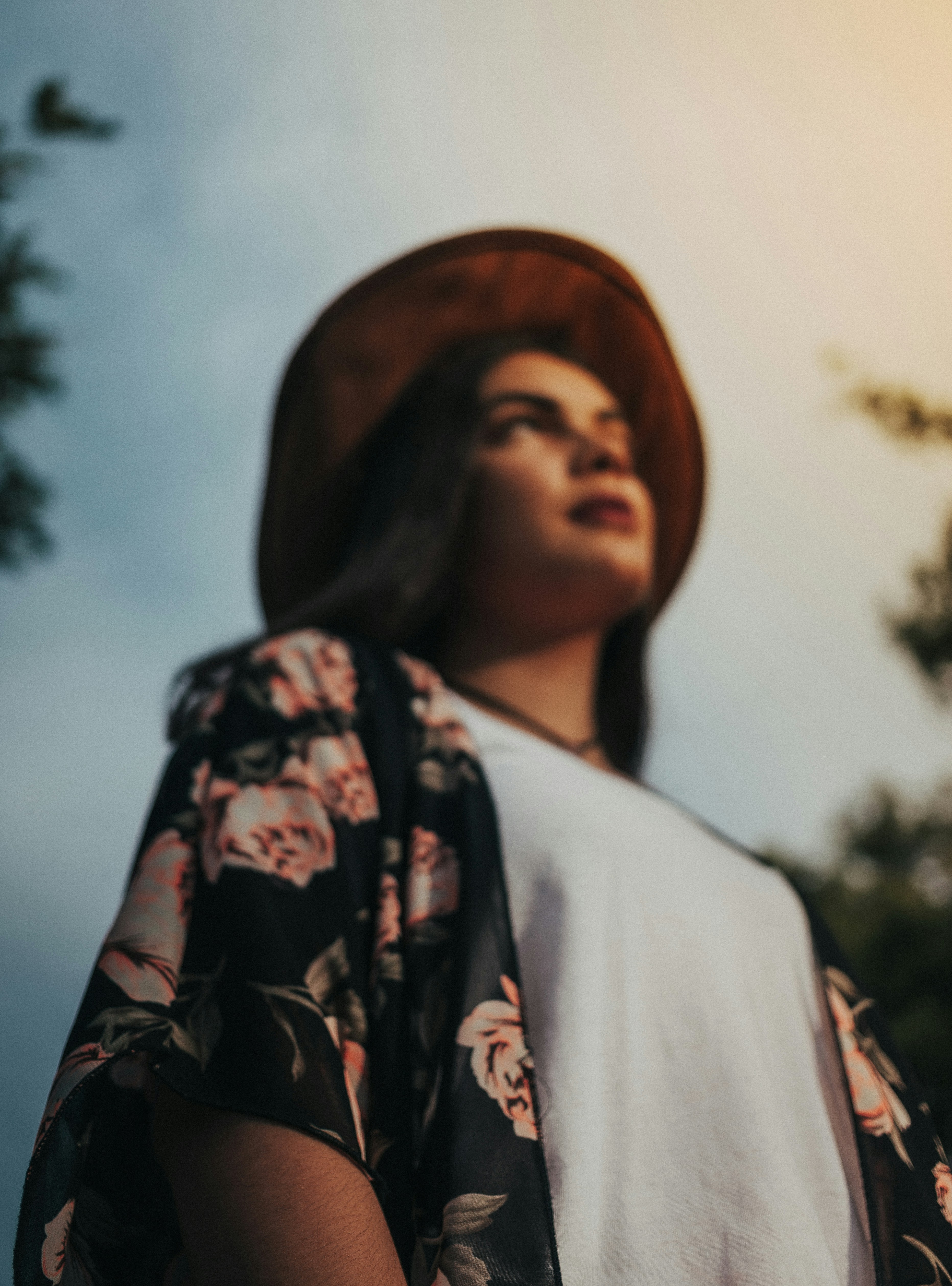 A young woman gazes upward, adorned in a floral kimono and hat, with a soft glow from the setting sun illuminating her features.