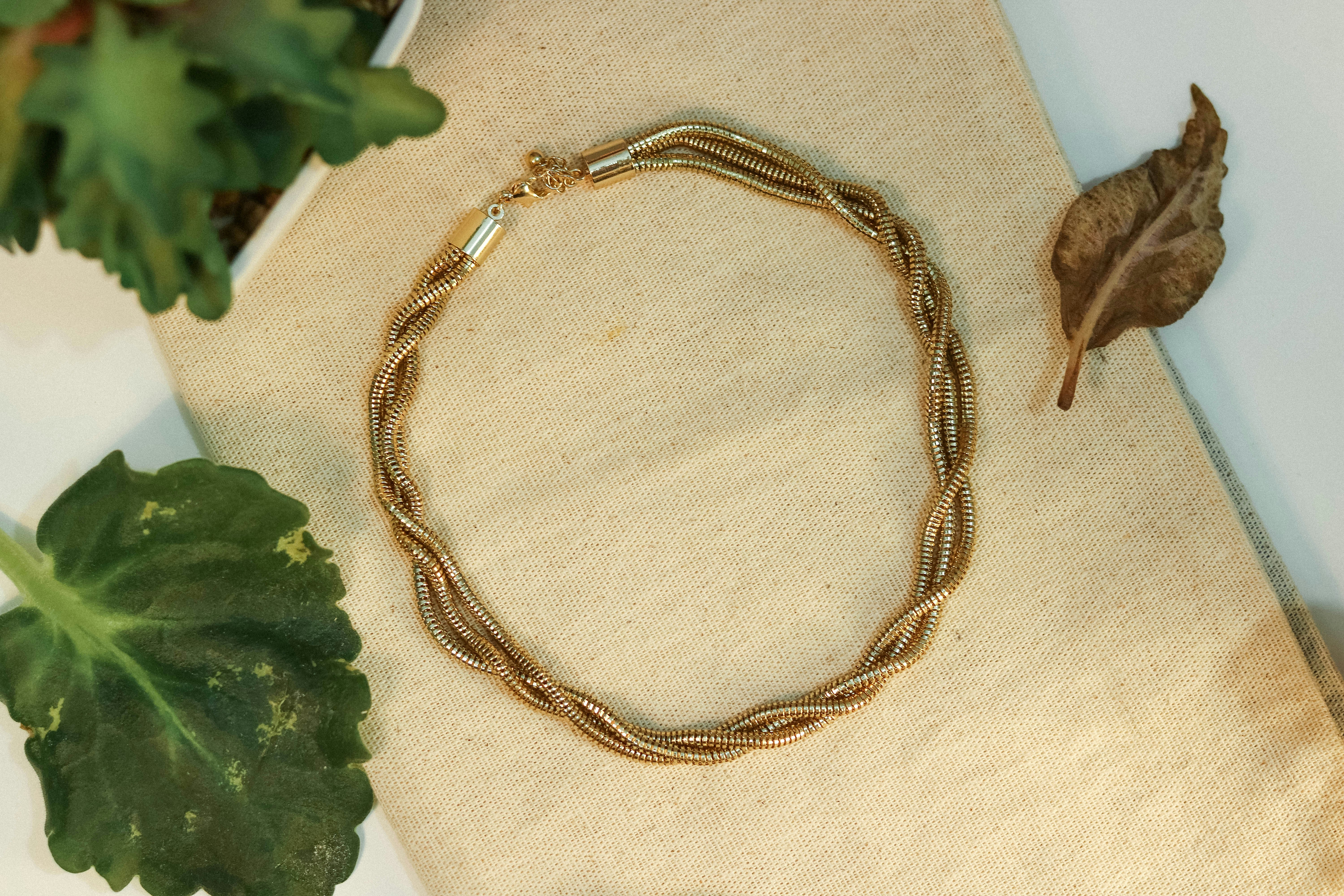 Intricately designed gold necklace resting on a textured backdrop, surrounded by greenery and natural elements.