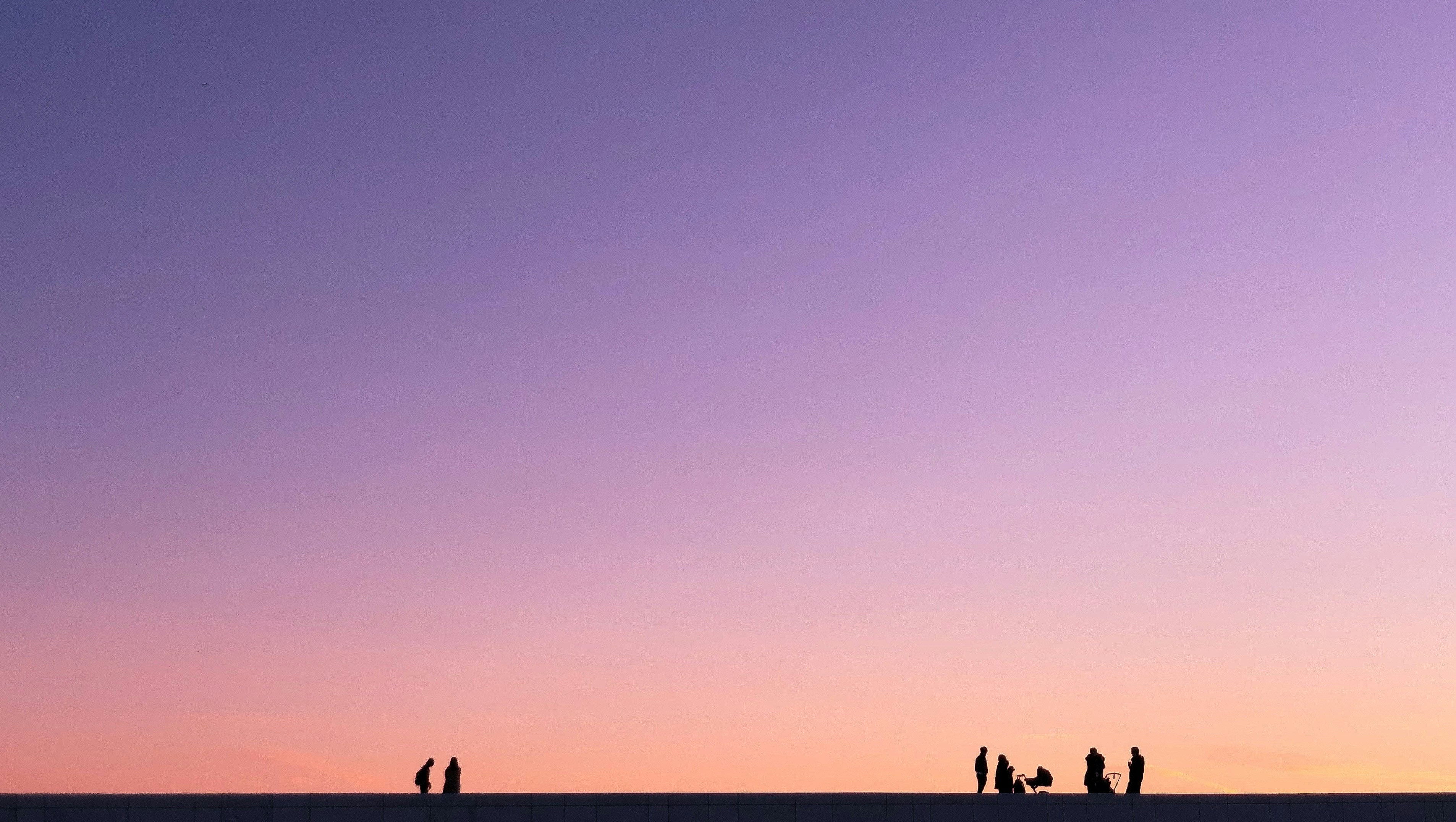 Silhouettes of small groups along a flat horizon at sunset, with a gradient sky shifting from purple to warm orange; the scene emphasizes vast sky and distant figures.