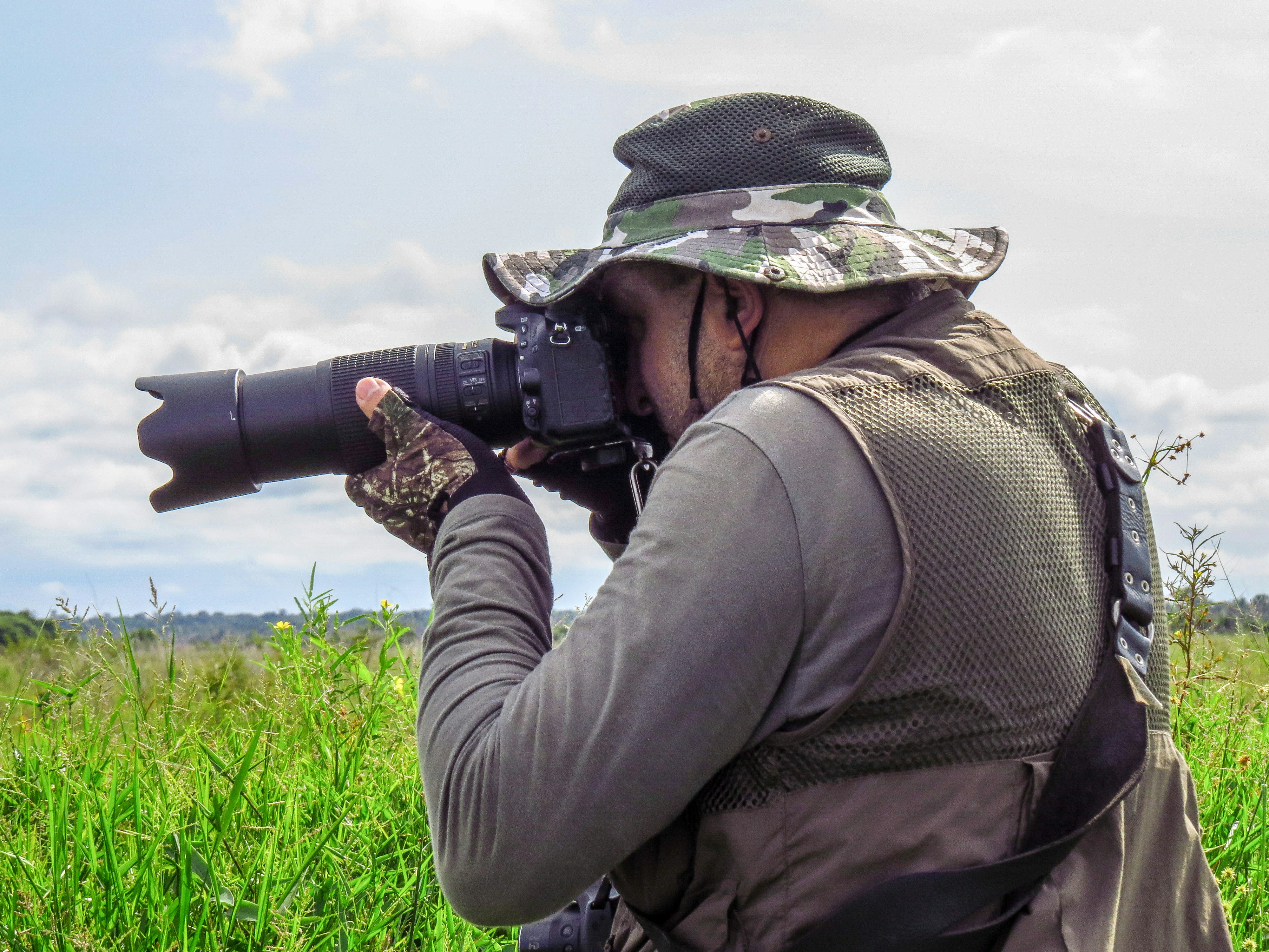 Photographer in camouflage attire aiming a DSLR camera amidst tall grass under a cloudy sky.