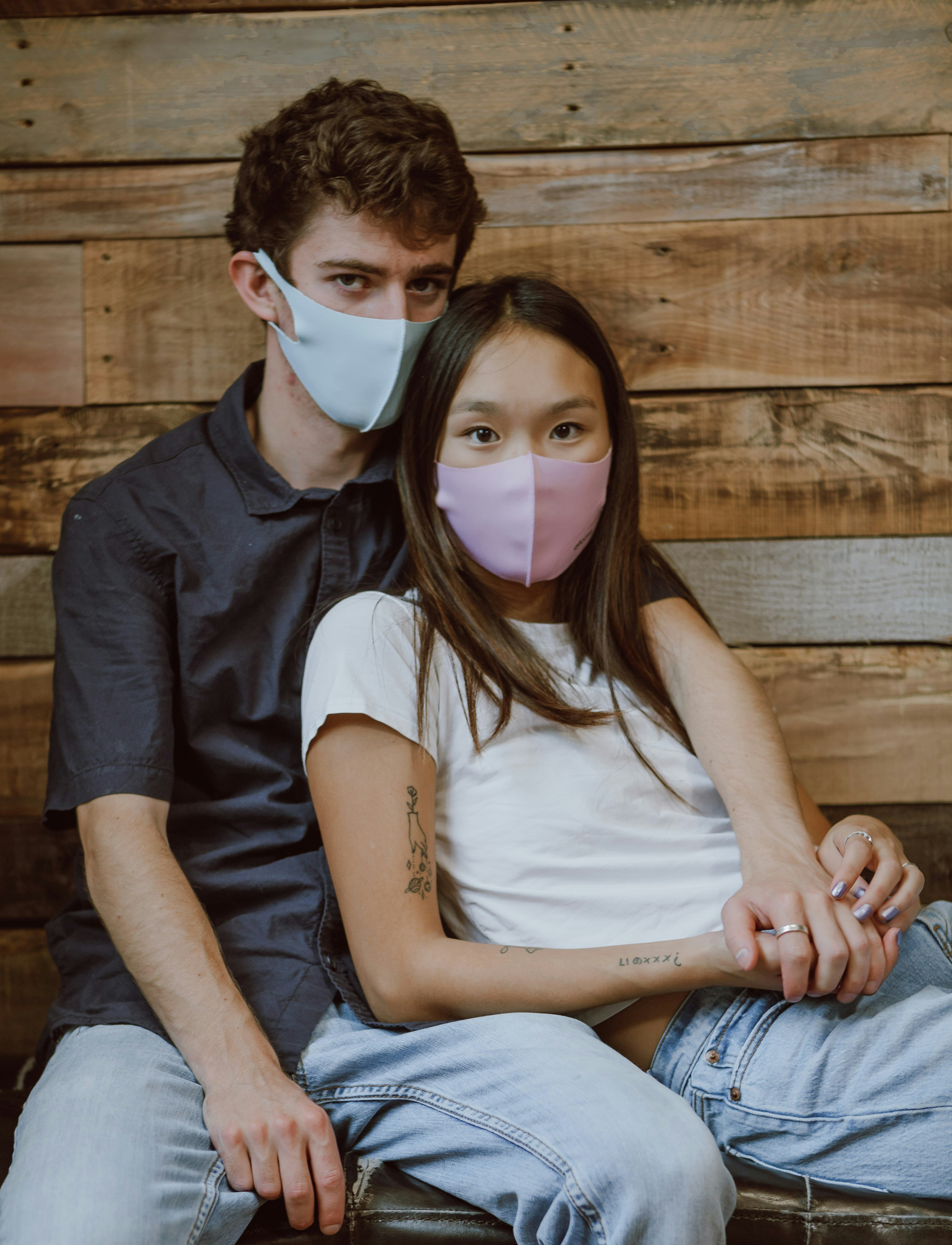 Couple wearing pastel-colored masks, seated against a rustic wooden backdrop, exuding a sense of connection and contemporary style.