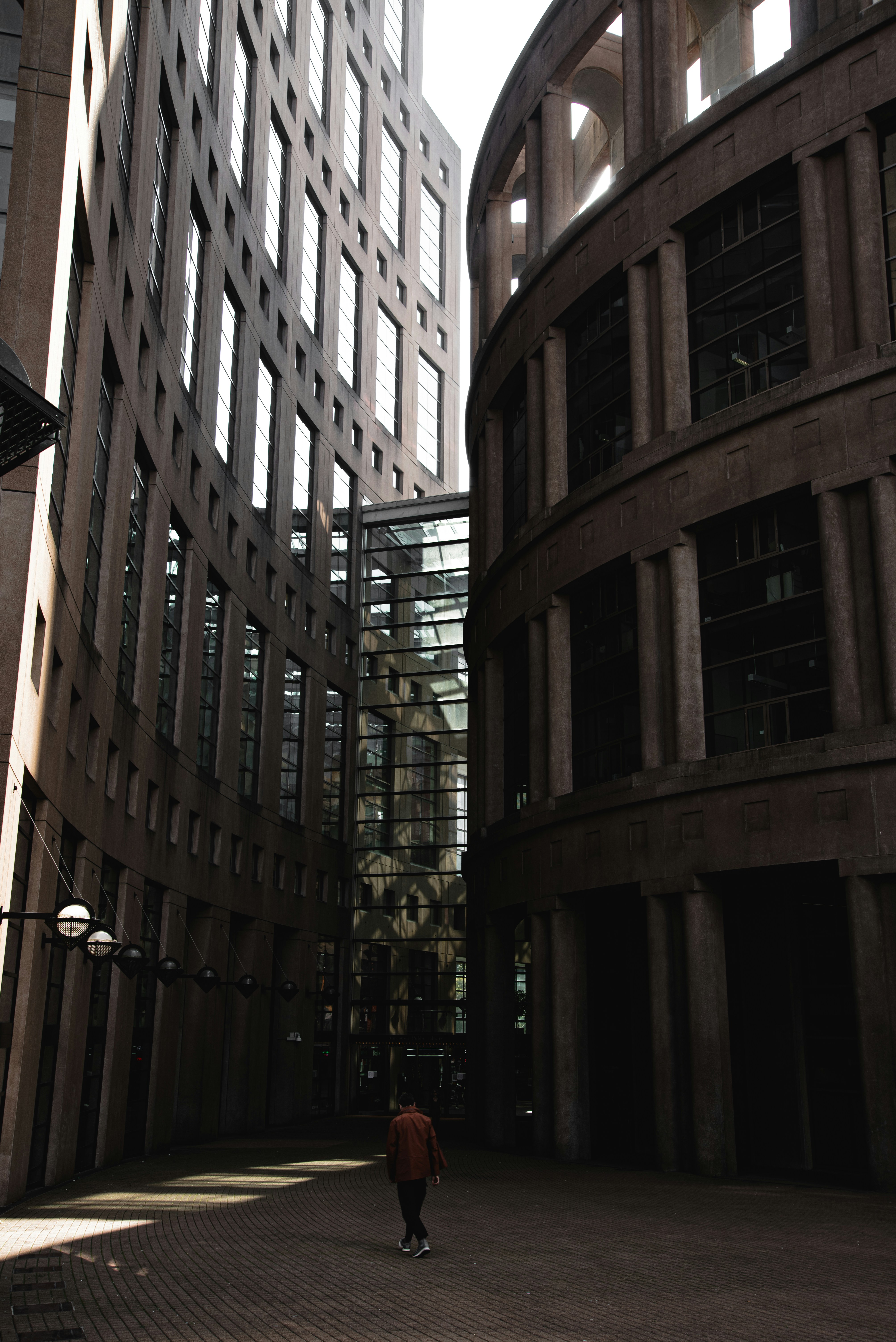 A solitary figure walks through a narrow urban passage framed by strikingly curved buildings and reflective glass surfaces.