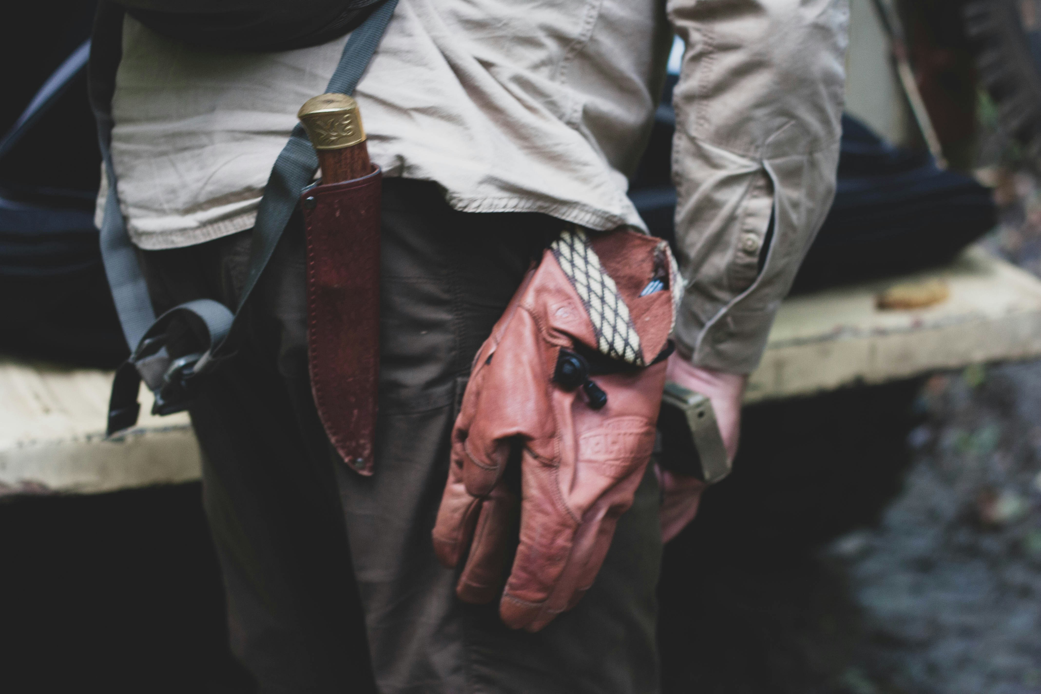 Close-up of a rugged explorer's gear, featuring a knife sheath and a weathered glove, set against a blurred outdoor backdrop.