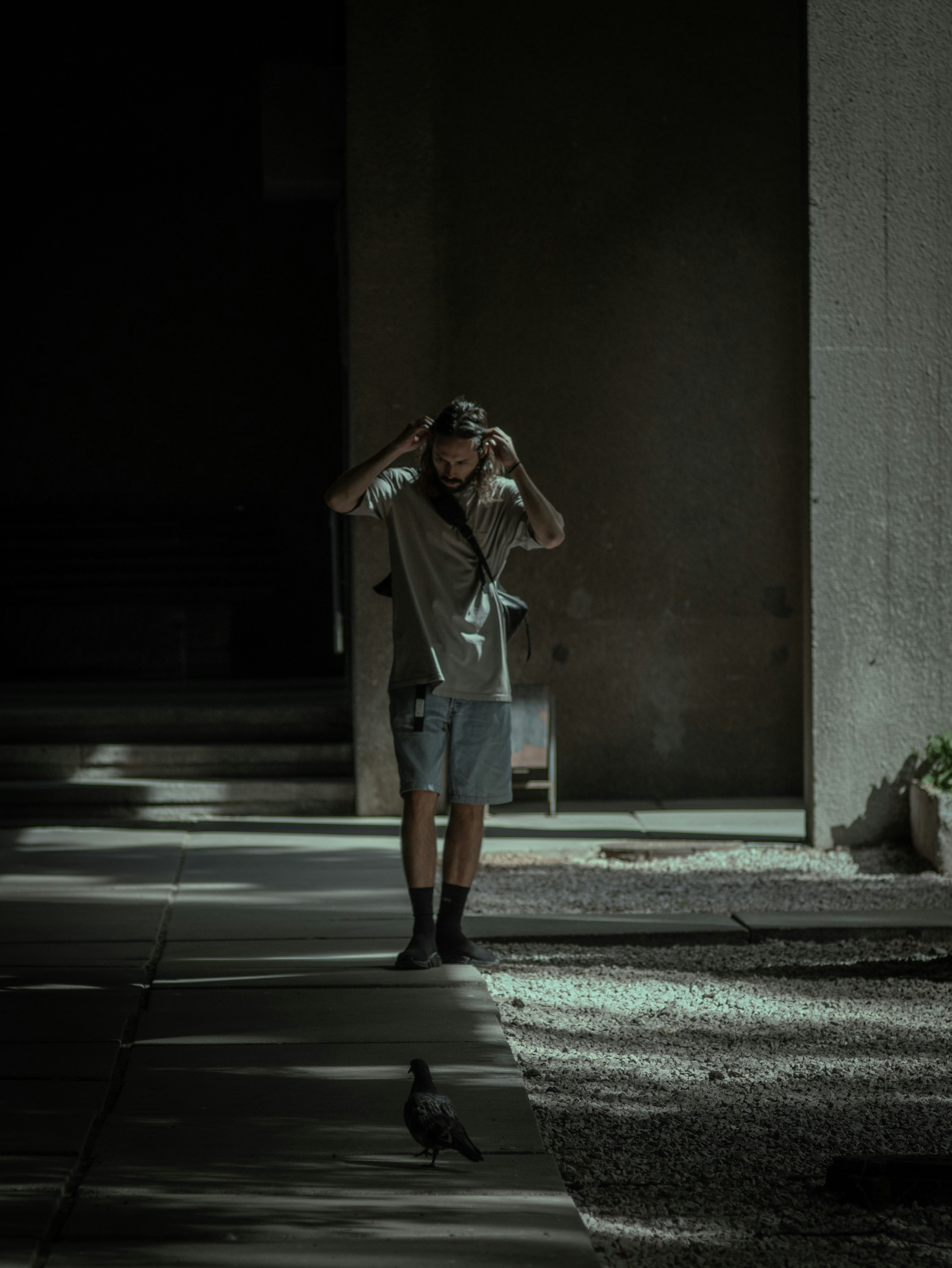 A man adjusts his headphones in a dimly lit urban space, while a bird walks nearby on the pavement.
