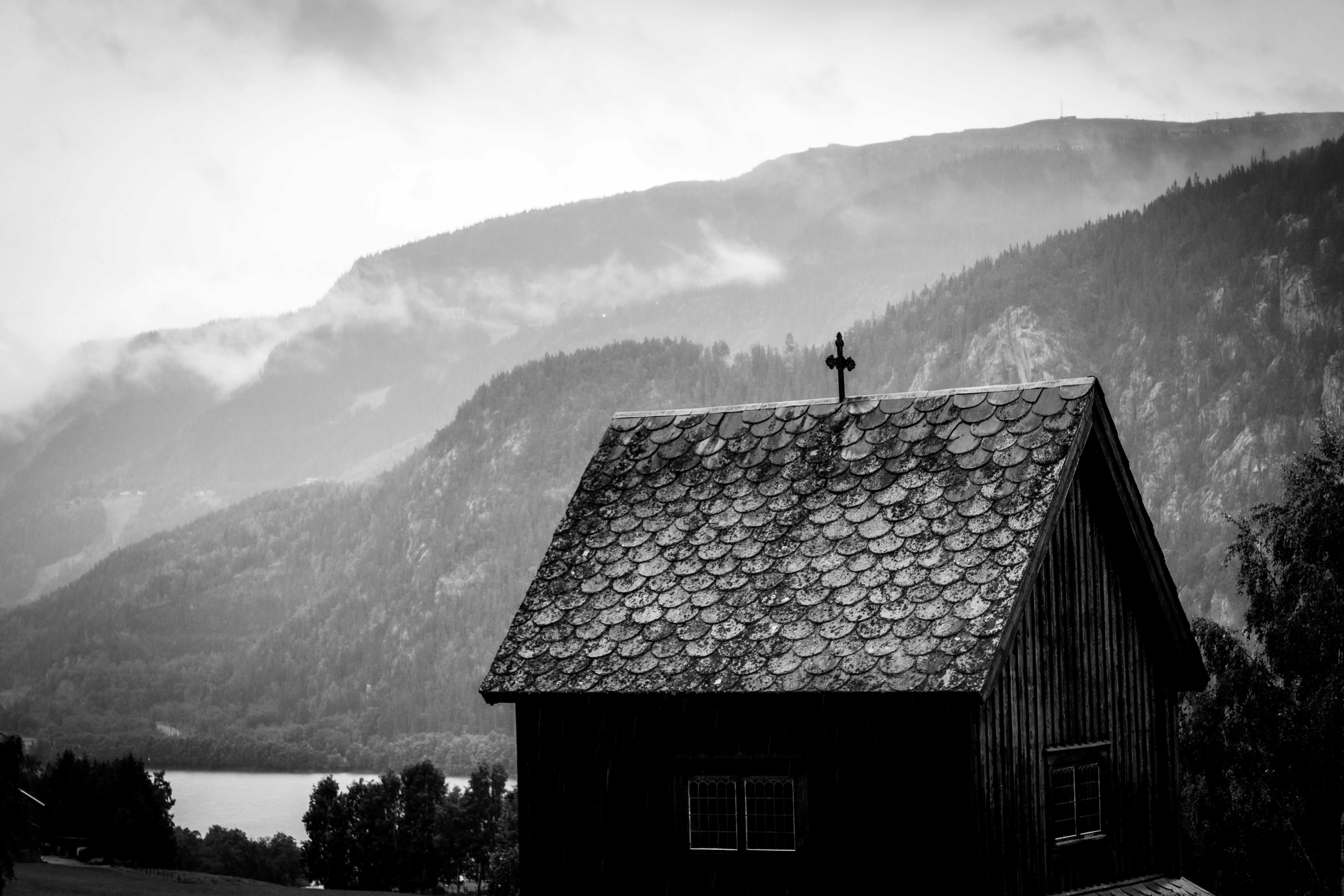 A quaint wooden cabin with a shingled roof stands against a backdrop of misty mountains, shrouded in monochrome tones. The scene evokes a sense of solitude and tranquility.