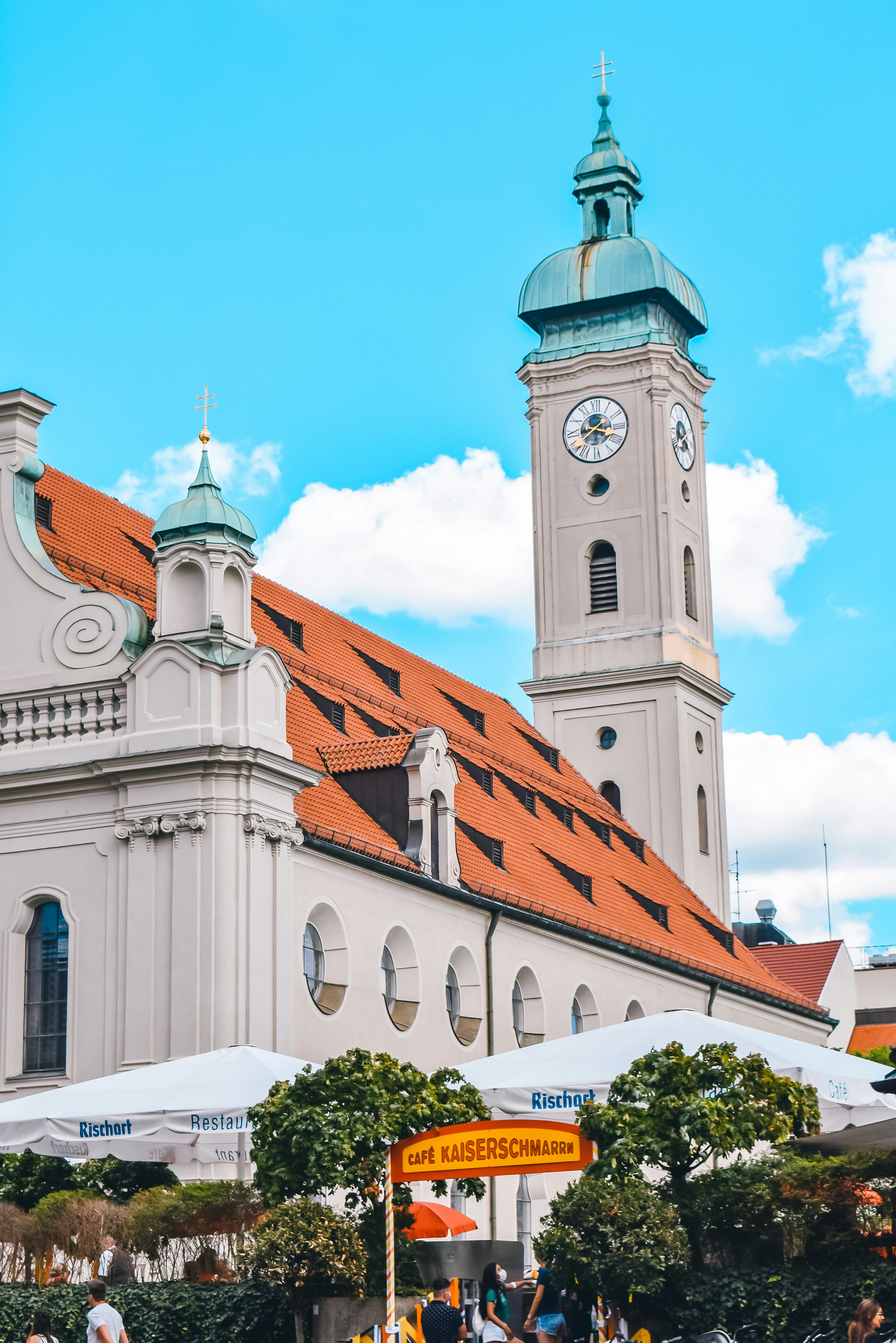 Historic building with a prominent clock tower, surrounded by outdoor café seating and lush greenery.