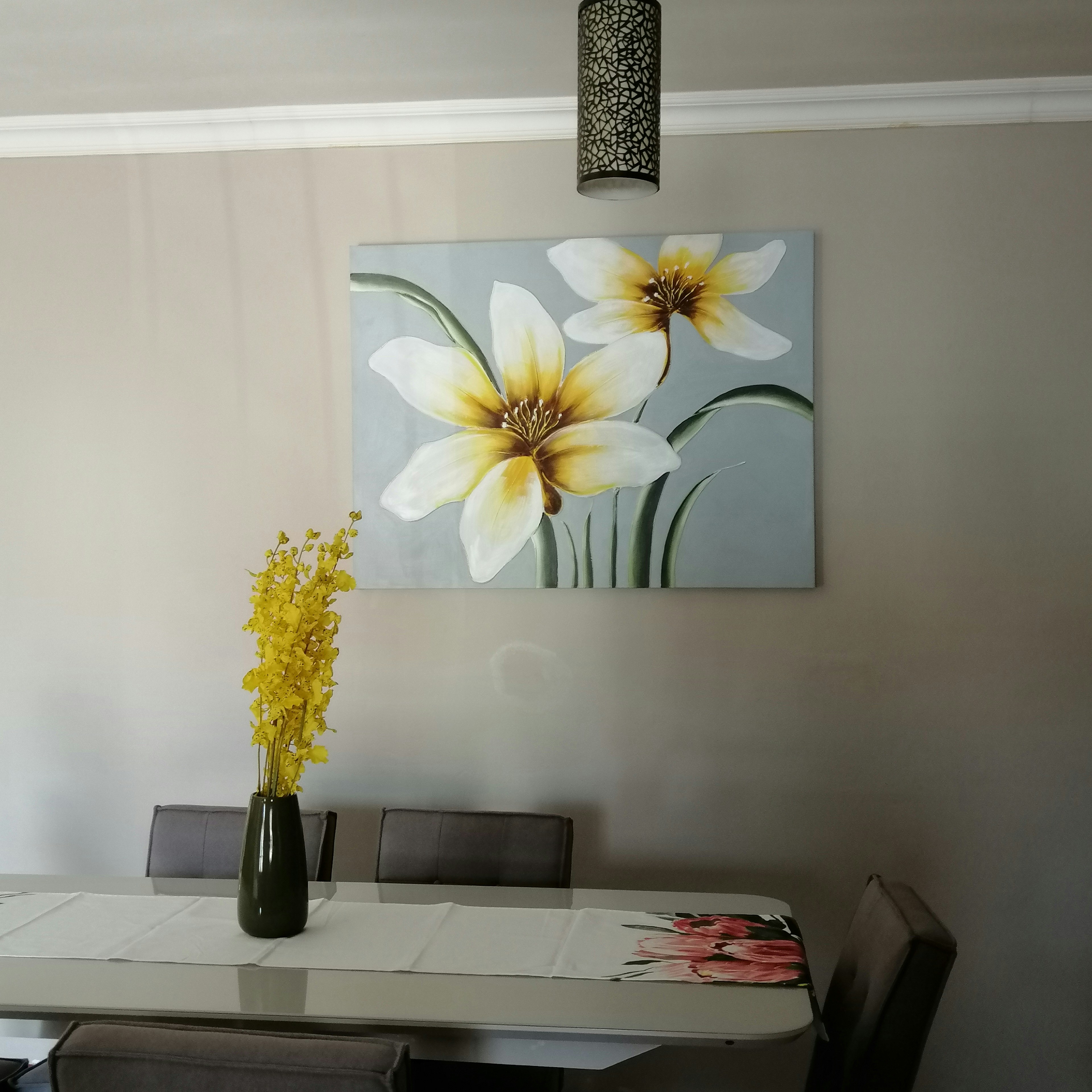 Canvas painting of white and yellow flowers hangs on a pale wall above a dining table. A vase of tall yellow blooms anchors the foreground, adding vertical contrast.
