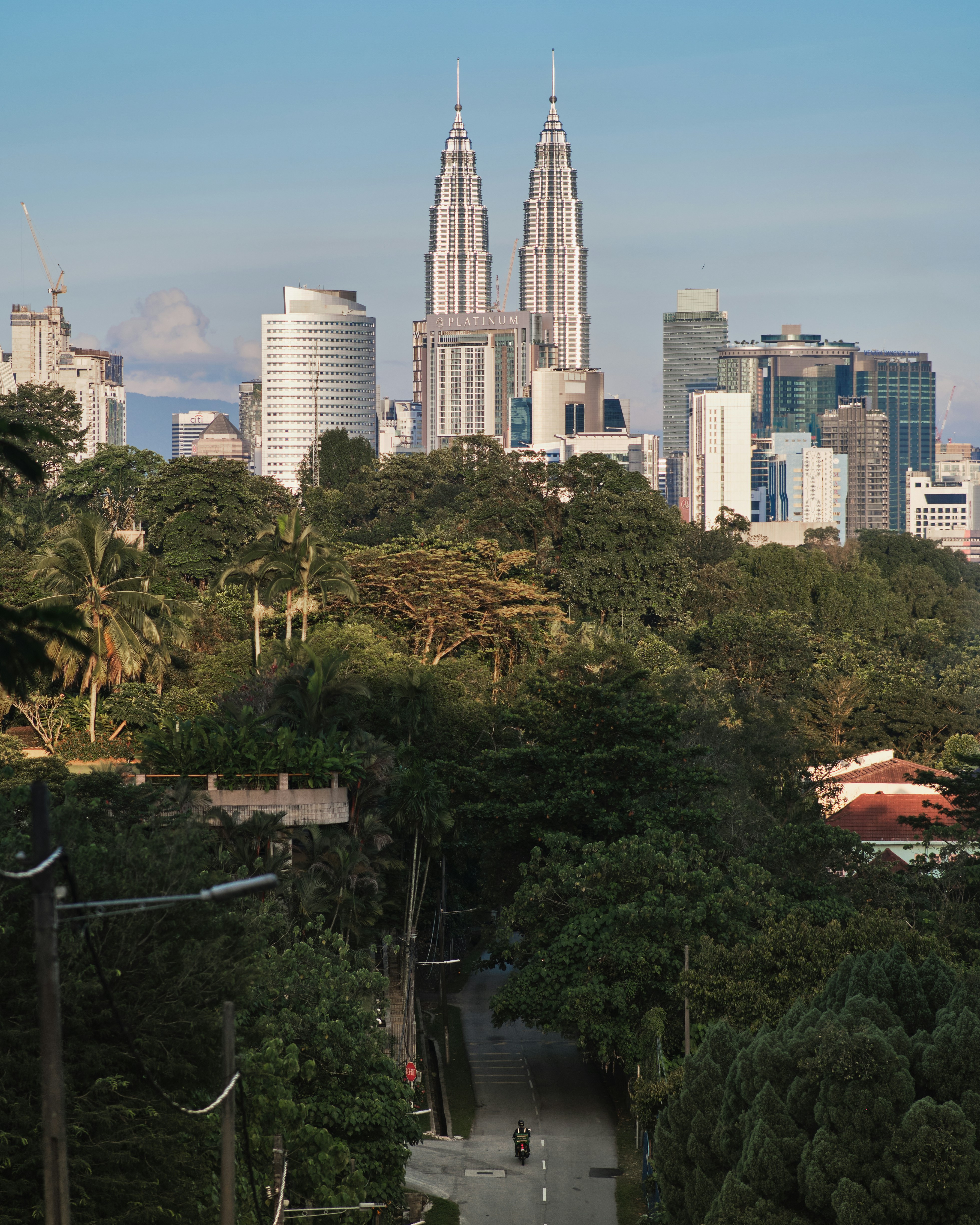 The iconic Petronas Towers rise above a lush canopy of trees, blending urban architecture with nature's beauty. A lone motorbike travels down a winding road, hinting at the city's vibrant life.