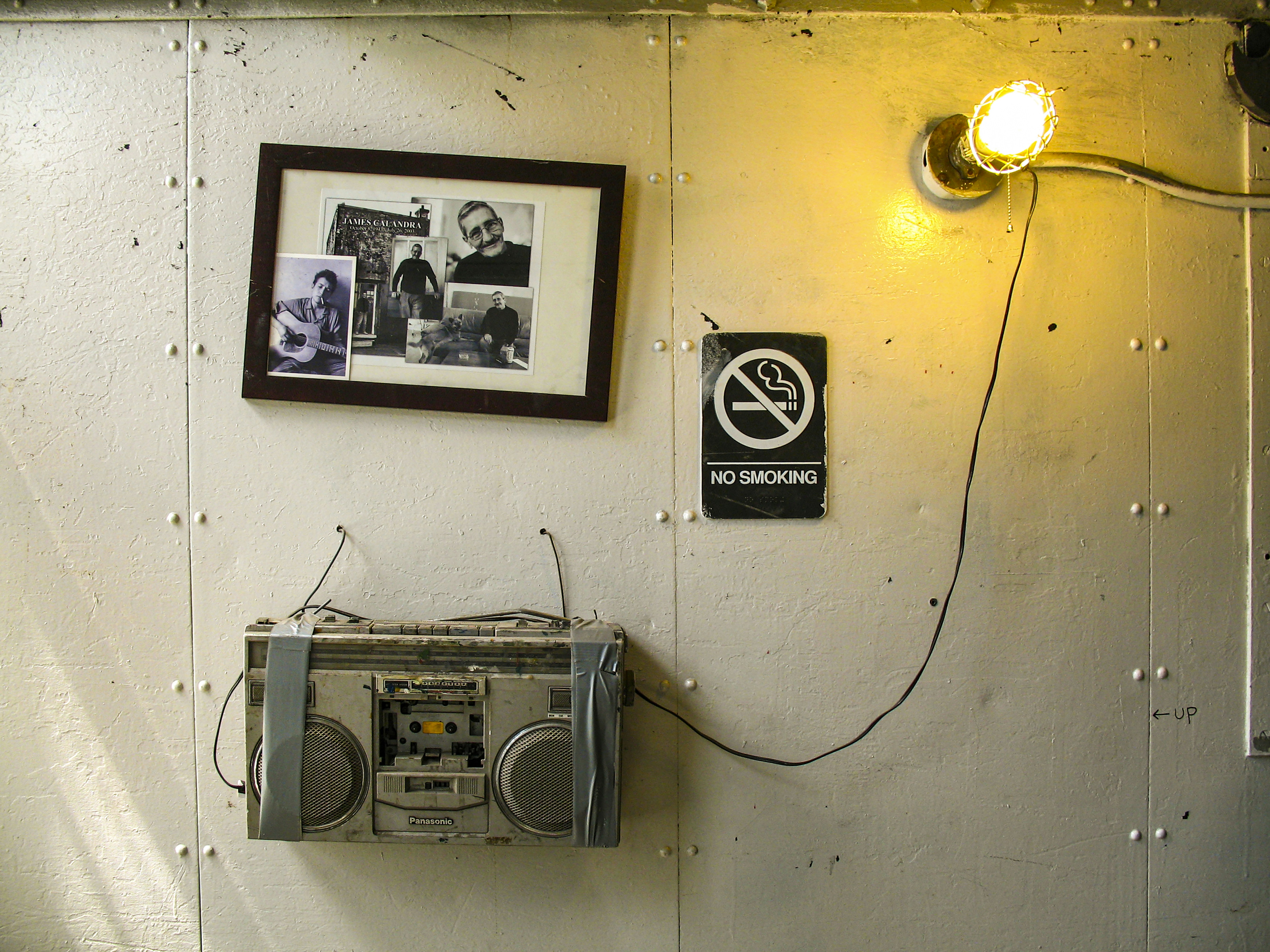 A vintage boombox rests on a riveted metal wall beside a framed photo collage and a No Smoking sign, lit by a single warm bulb.
