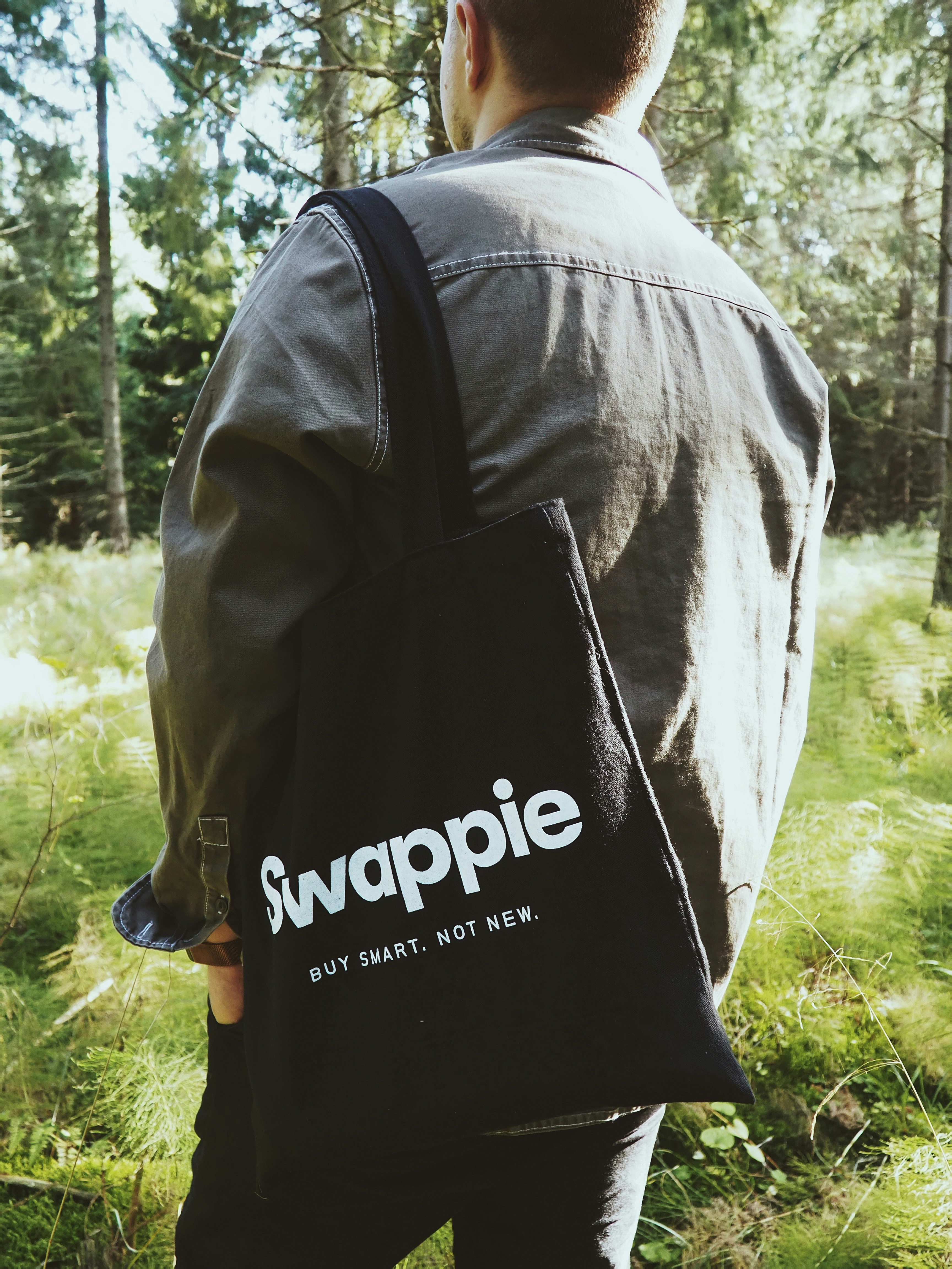 A person with a Swappie tote slung over the shoulder stands in a sunlit forest, with the Swappie logo clearly visible on the bag.