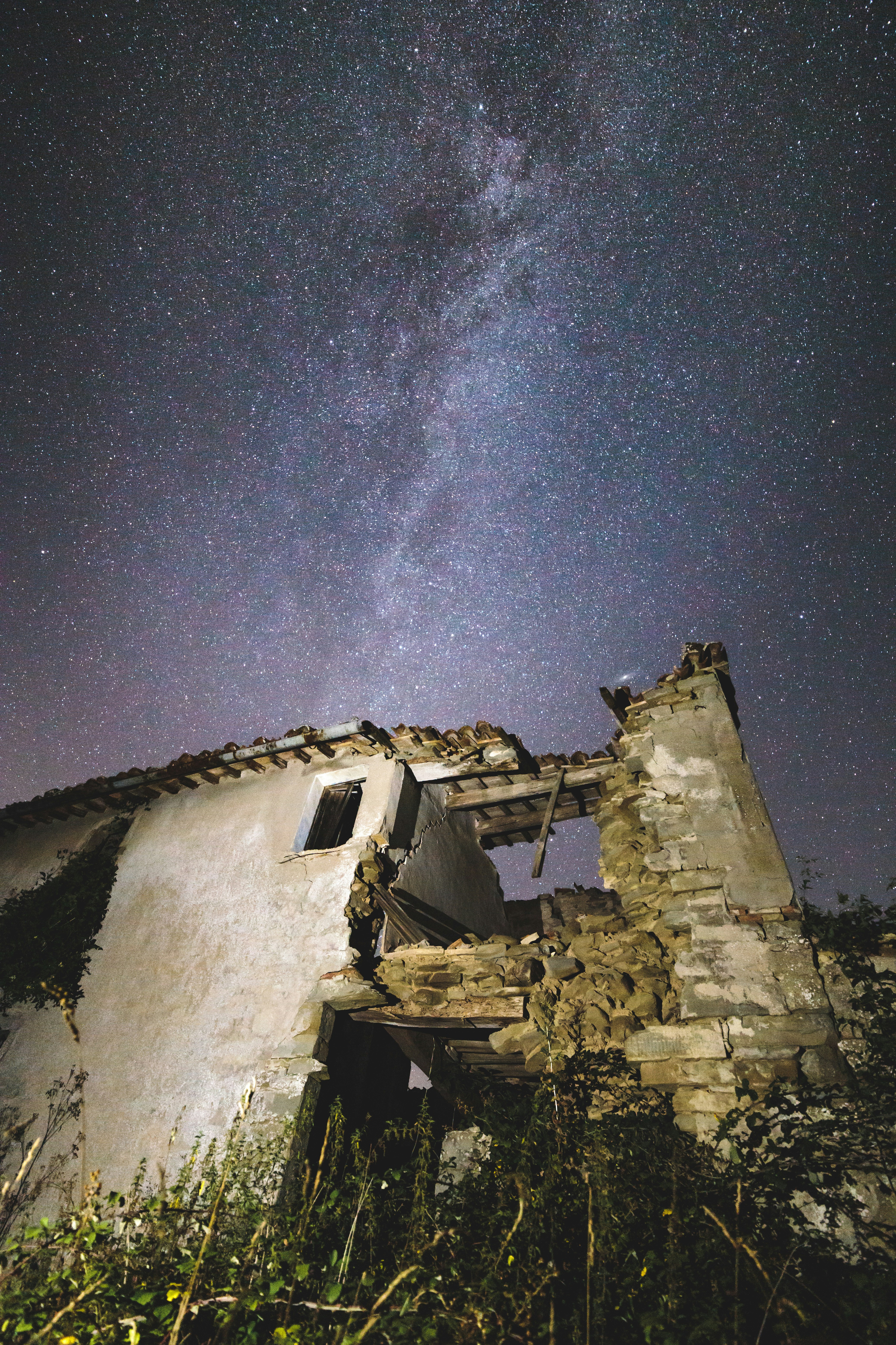 Abandoned house with crumbling walls under a vibrant night sky filled with stars and the Milky Way. The structure is overgrown with vegetation, adding to its eerie charm.