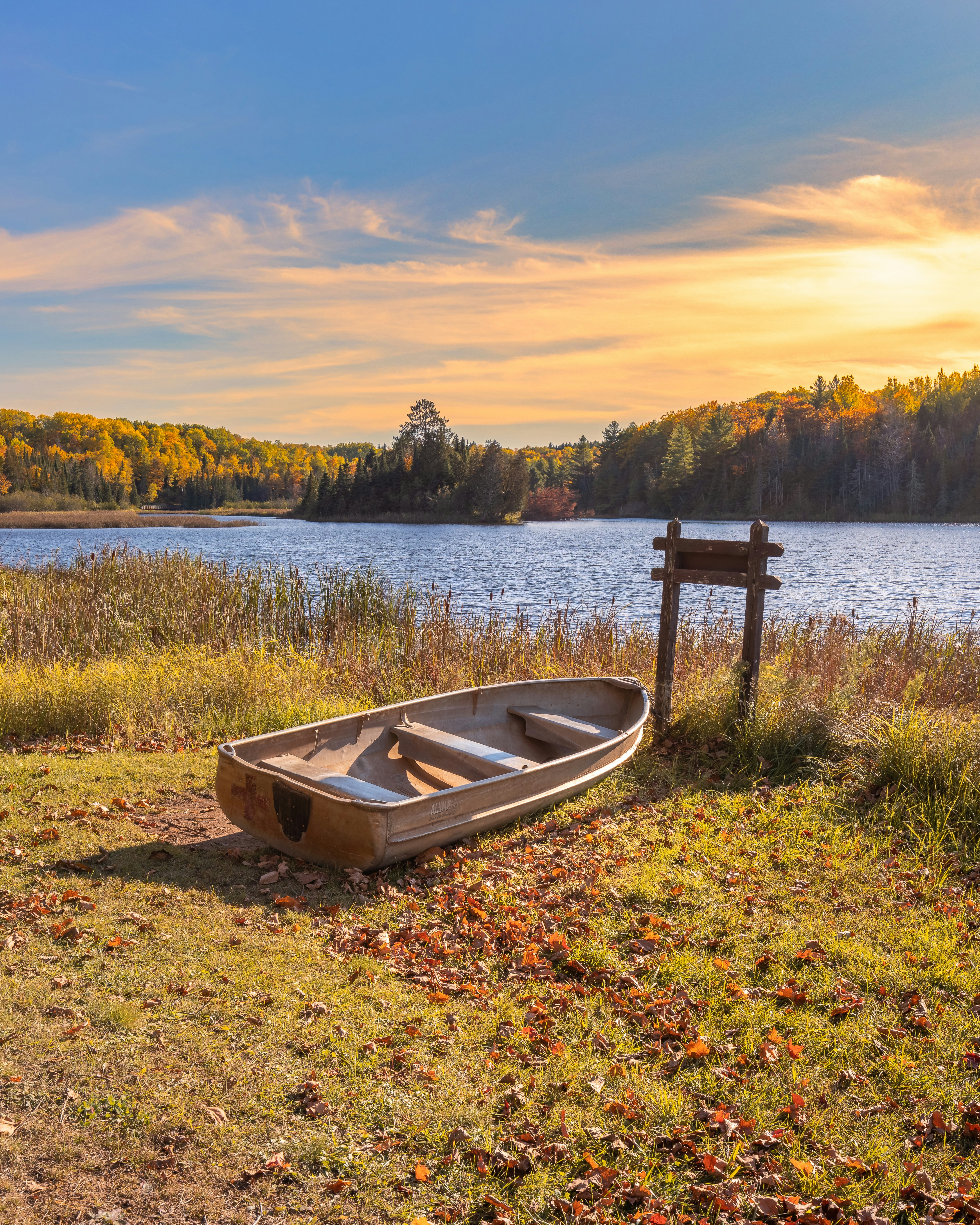 A rustic wooden boat rests on the shore, surrounded by vibrant autumn foliage and a tranquil lake. The warm hues of the sunset reflect on the water's surface.