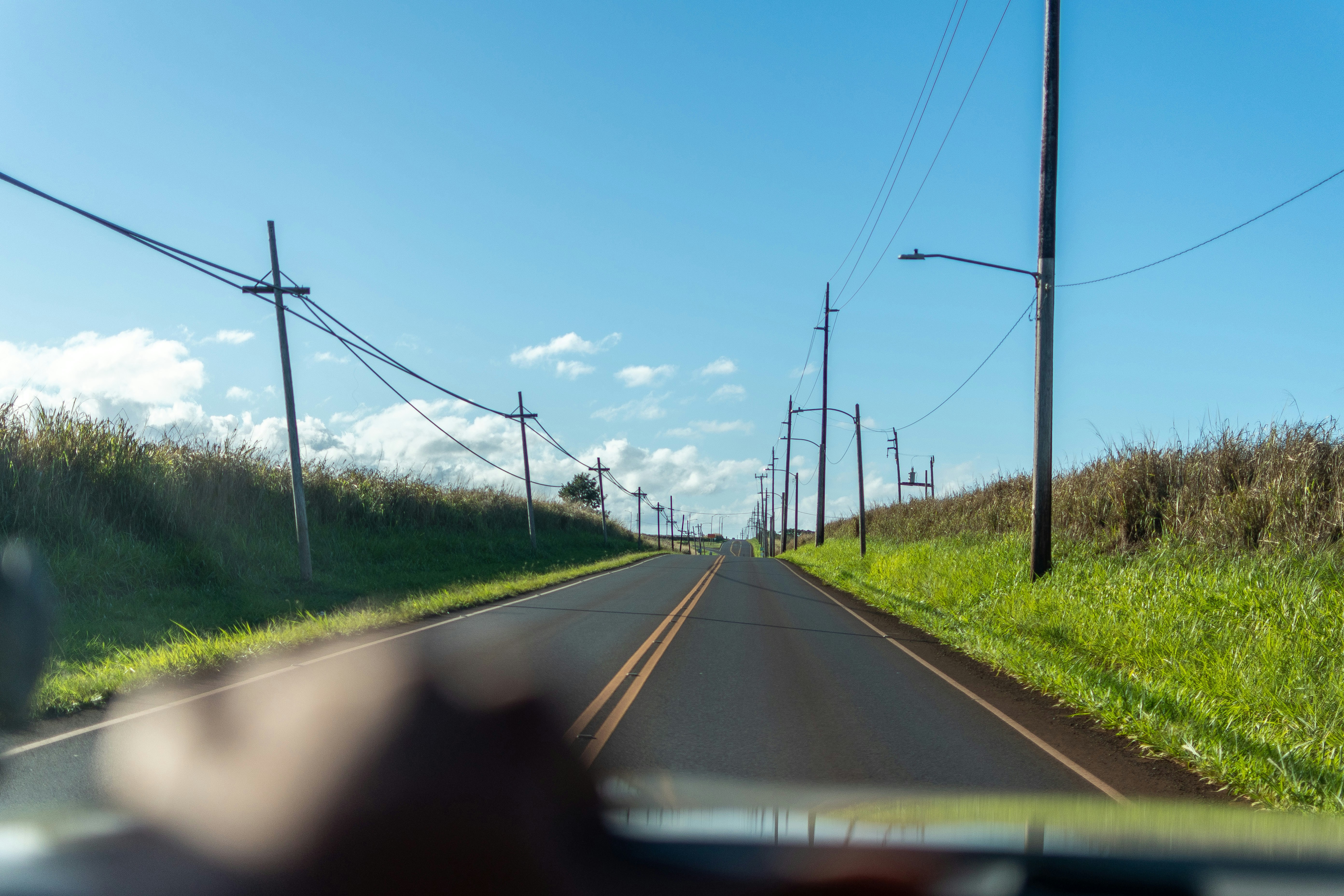 Straight road flanked by green fields under a clear blue sky, viewed from a vehicle's interior.