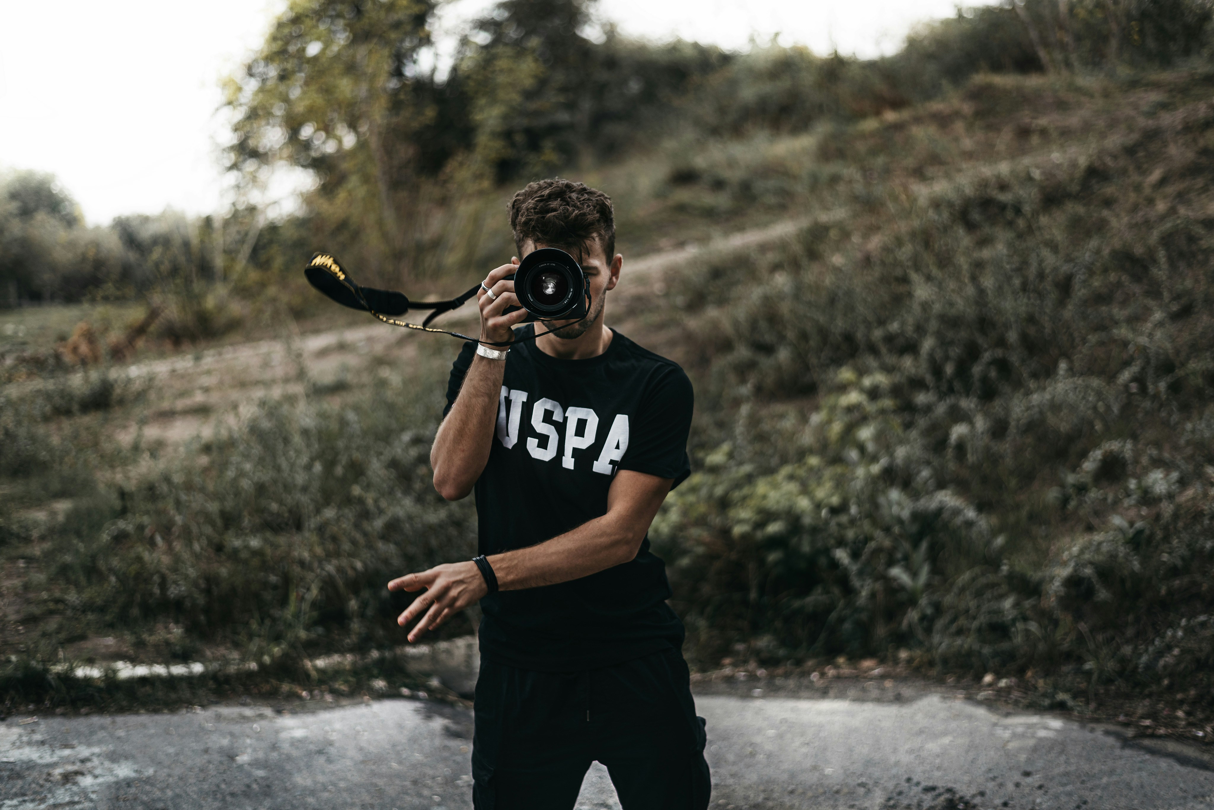 Photographer in a black T-shirt holds a camera in a grassy outdoor setting.