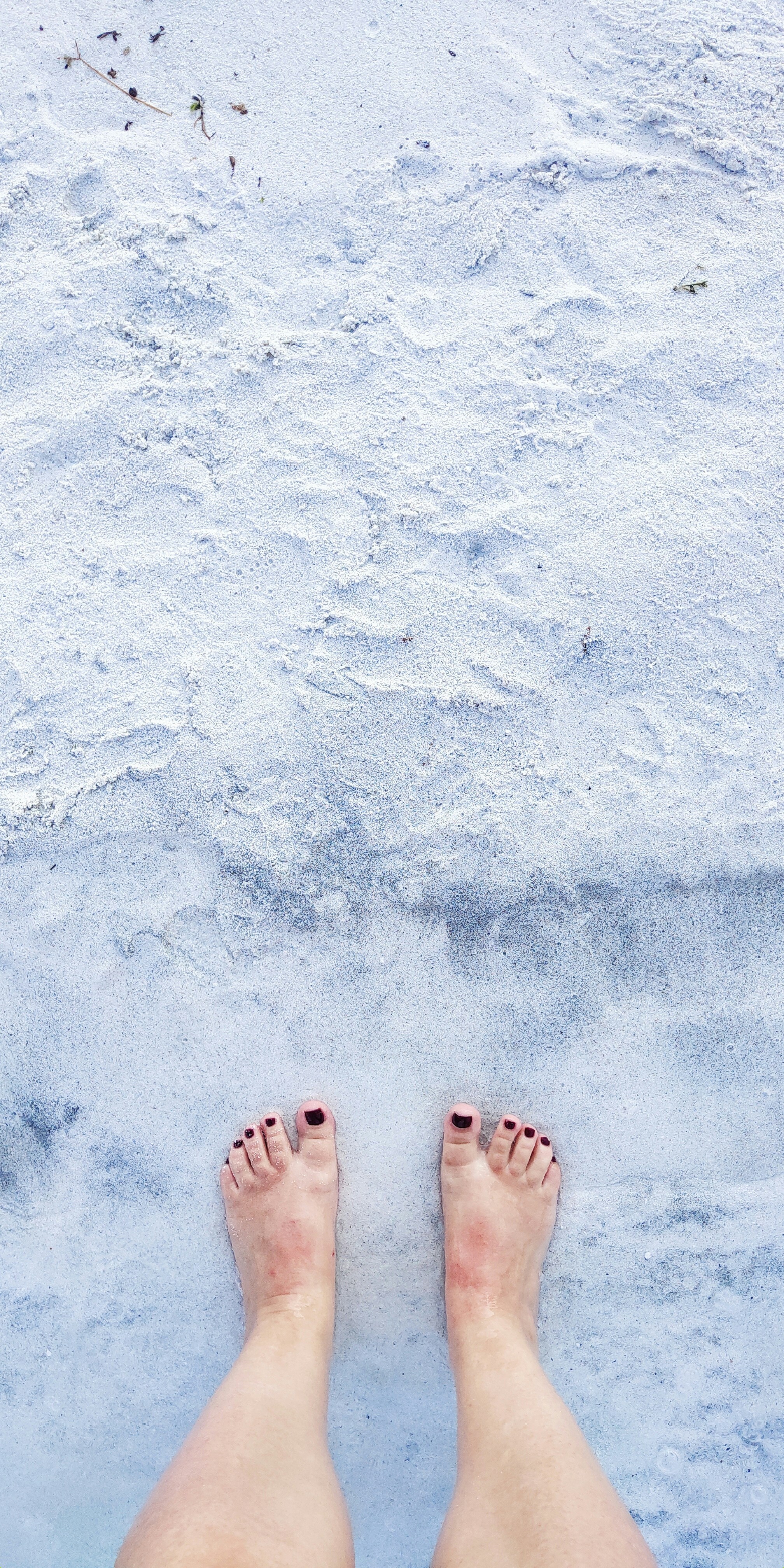 Photograph of bare feet standing on a pale blue snow-ice surface, toenails painted dark. The composition emphasizes winter texture and a moment of stillness.