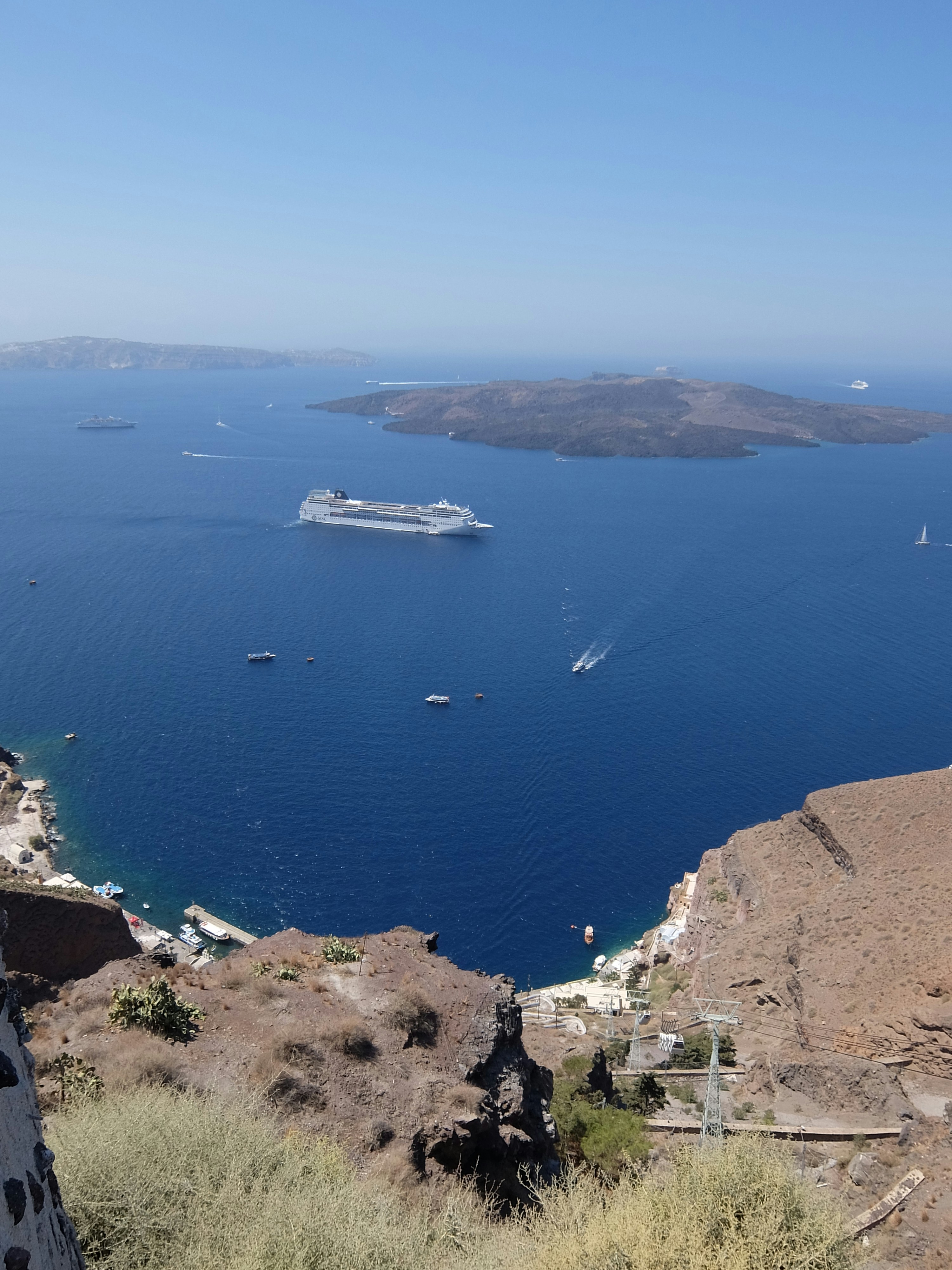 Sunlit caldera coastline photographed from above, featuring a large cruise ship and smaller boats gliding over the blue Aegean with rugged cliffs in the foreground.